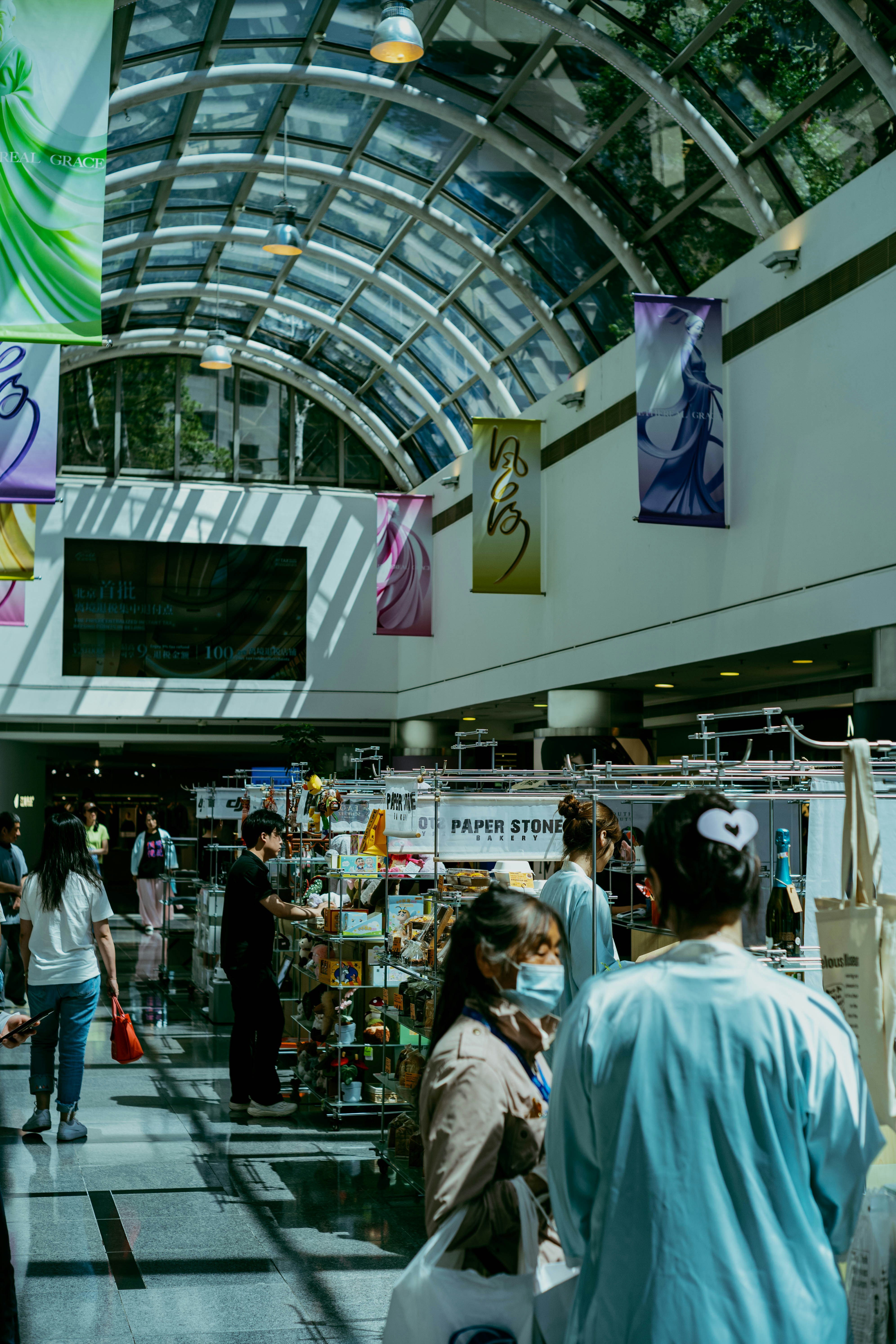 A bustling indoor market filled with vendors and shoppers, illuminated by natural light streaming through a glass ceiling. Colorful banners hang above, adding to the lively atmosphere.