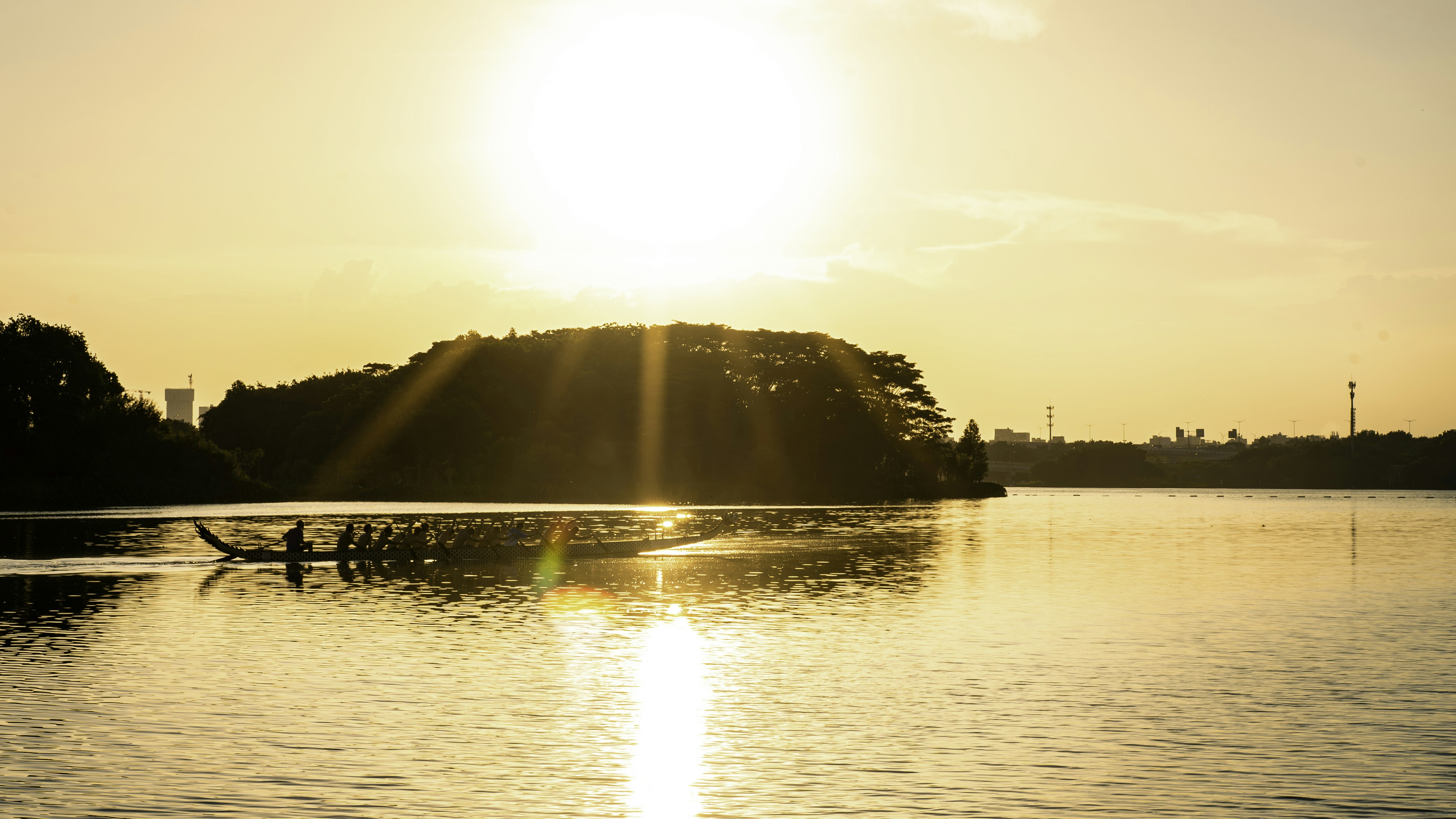 A dragon boat team is perfectly silhouetted against a brilliant golden sunset, their paddles moving in sync as they glide across the calm water. The sun, bursting from behind a tree-lined shore, casts dramatic rays of light and a shimmering reflection that leads the eye through the frame. This powerful image captures the dedication and synergy of teamwork amidst the tranquil beauty of the golden hour. | Sunset over a calm lake with a boat