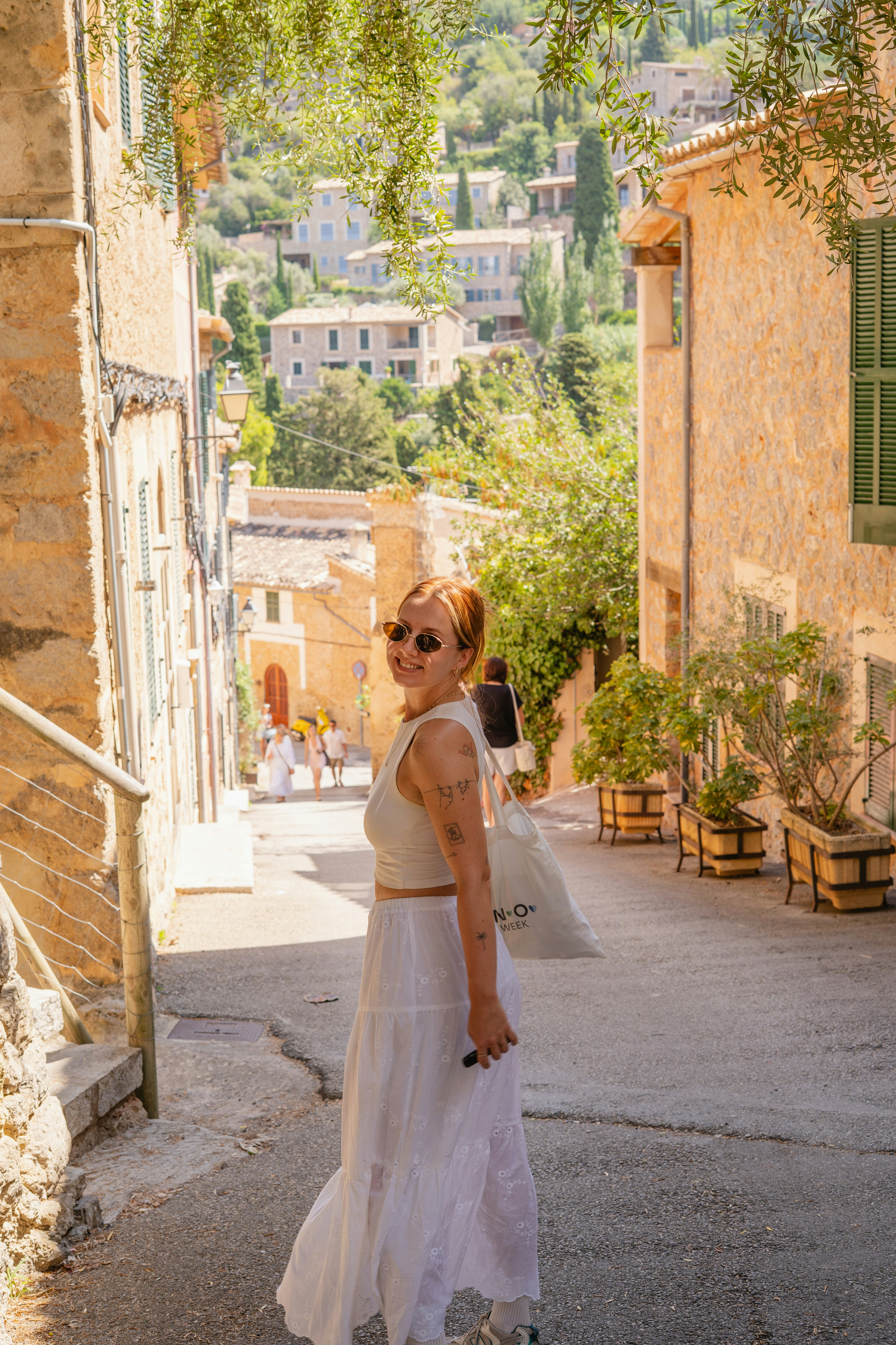 A woman in a flowing white dress walks down a charming stone pathway, surrounded by picturesque buildings and lush greenery.