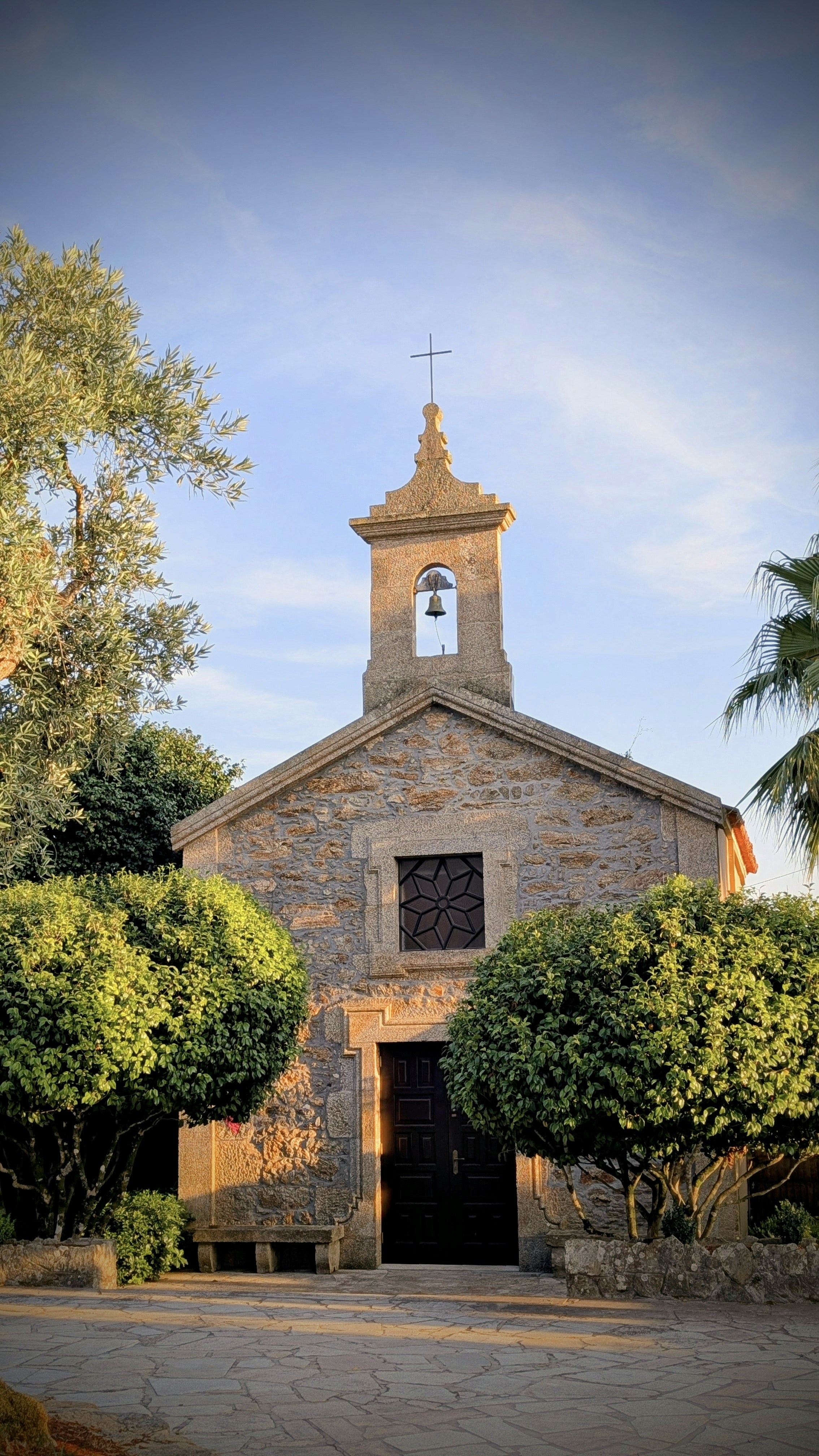 A historic church stands against the warm colors of sunset, its silhouette glowing in the evening sky. The blend of architecture and natural light creates a peaceful and timeless atmosphere. | Stone chapel with bell tower and trees at sunset