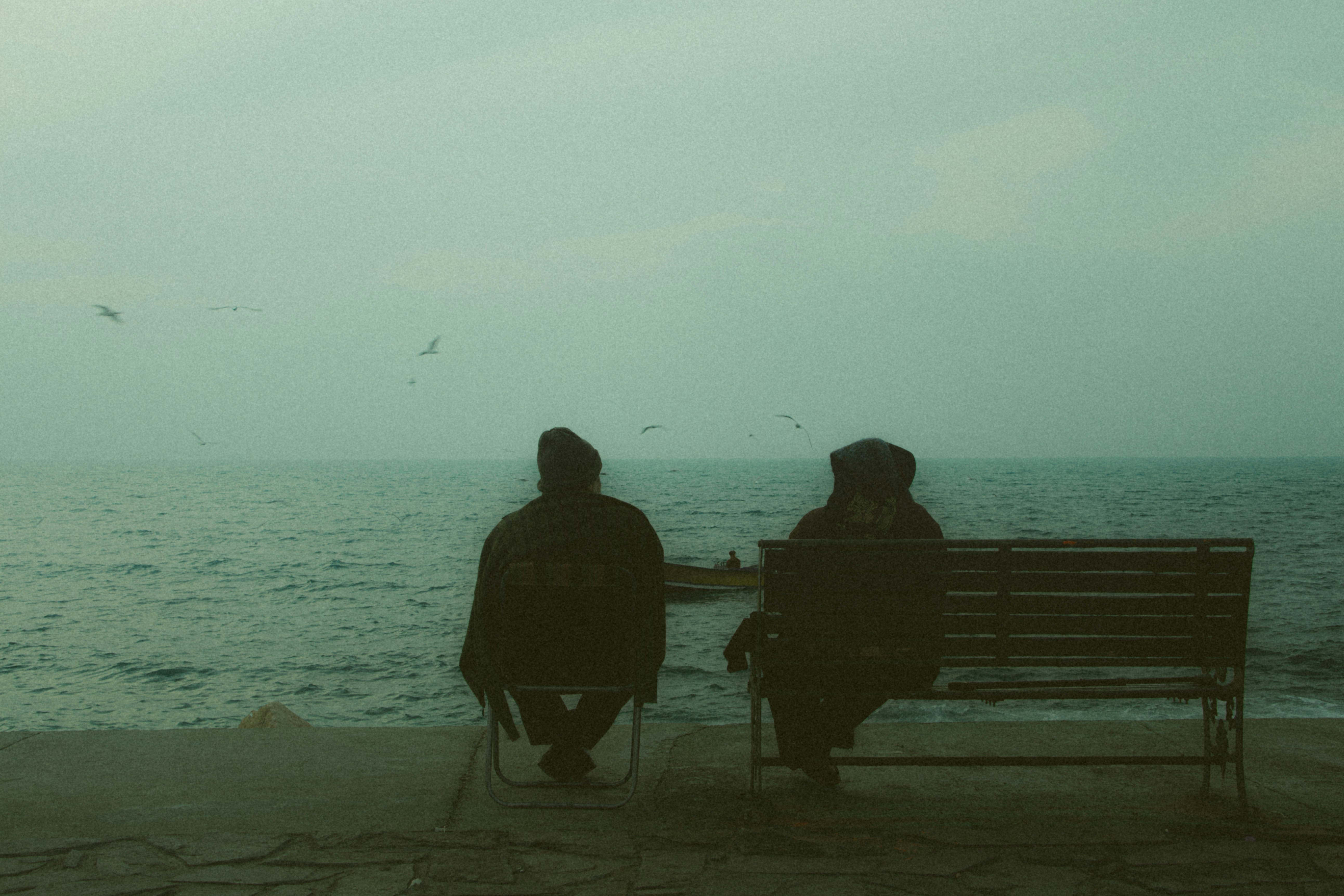 Two figures sit by the sea under a cloudy sky.
