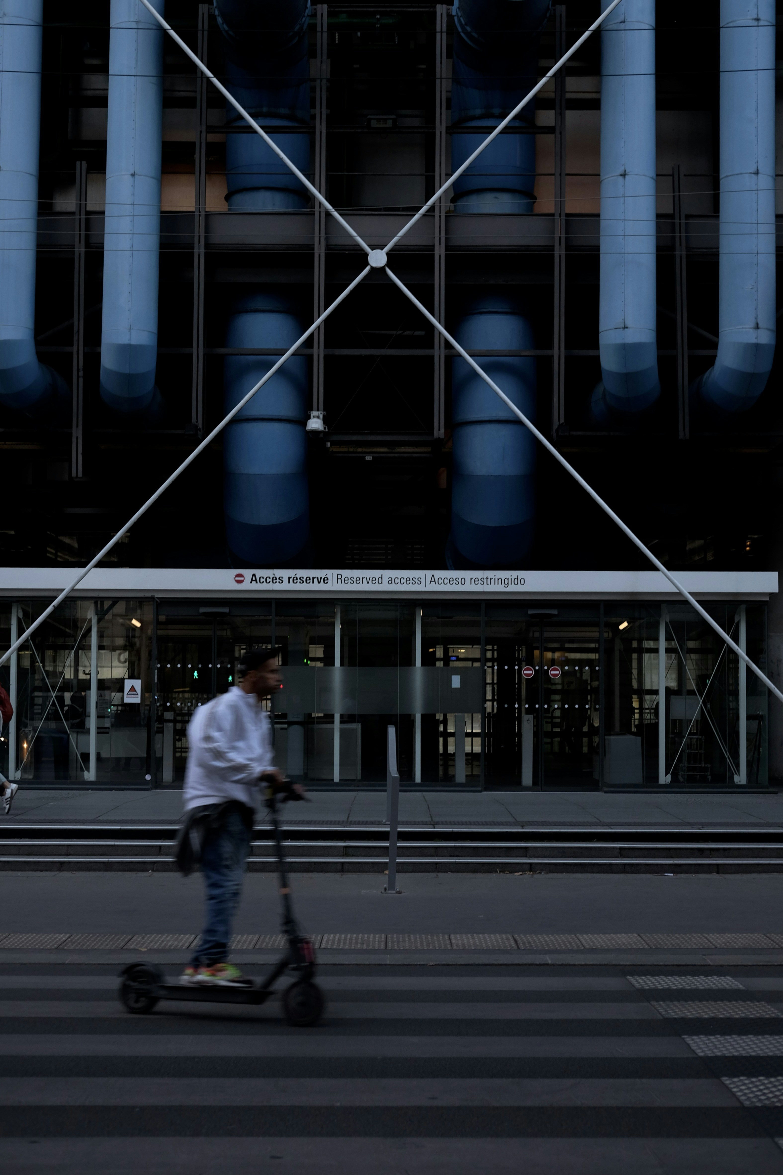 A person riding a scooter in front of a modern building with prominent blue pipes and a reserved access sign.