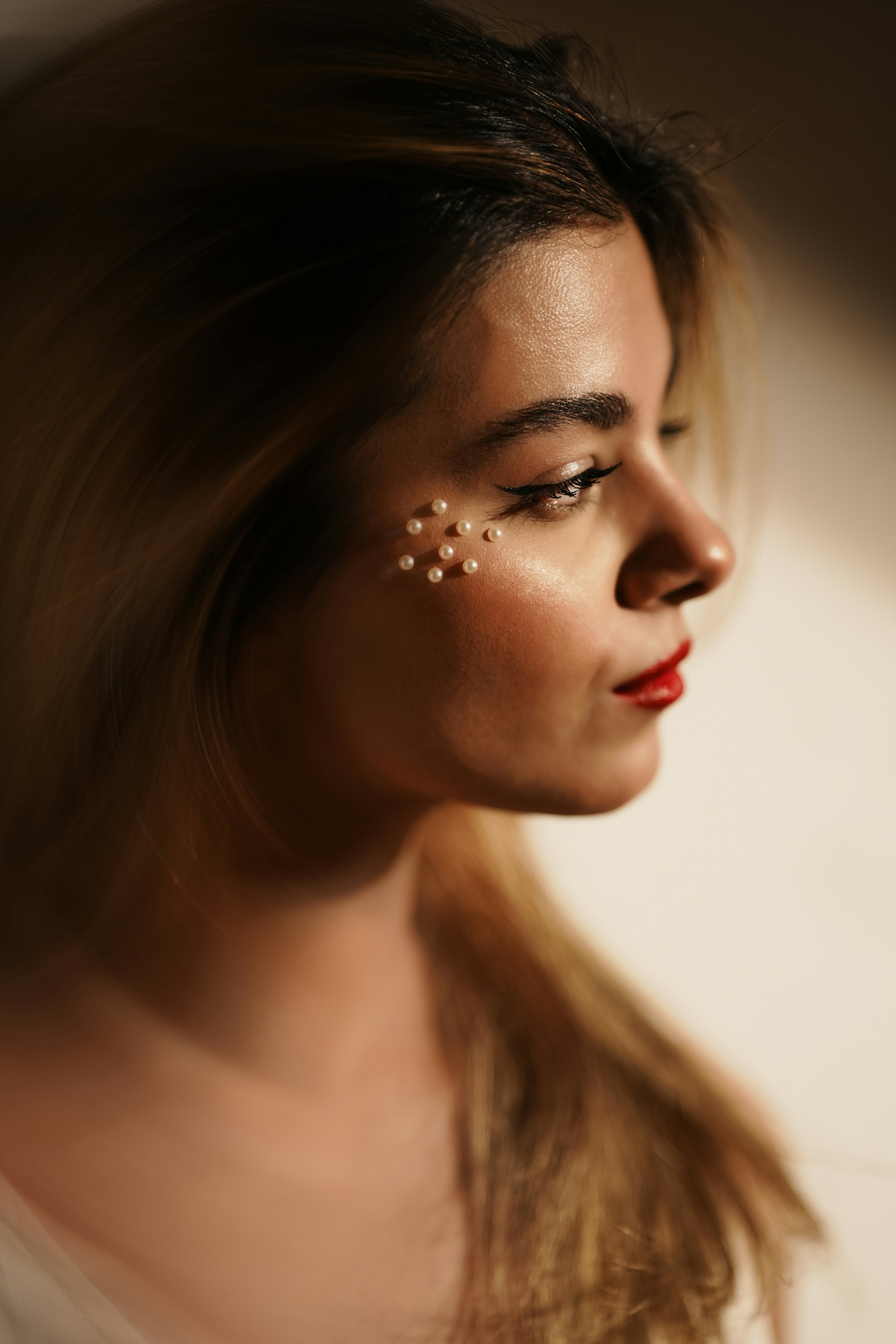 Profile view of a woman adorned with pearl-like embellishments on her cheek, showcasing a striking red lip against a softly lit background.