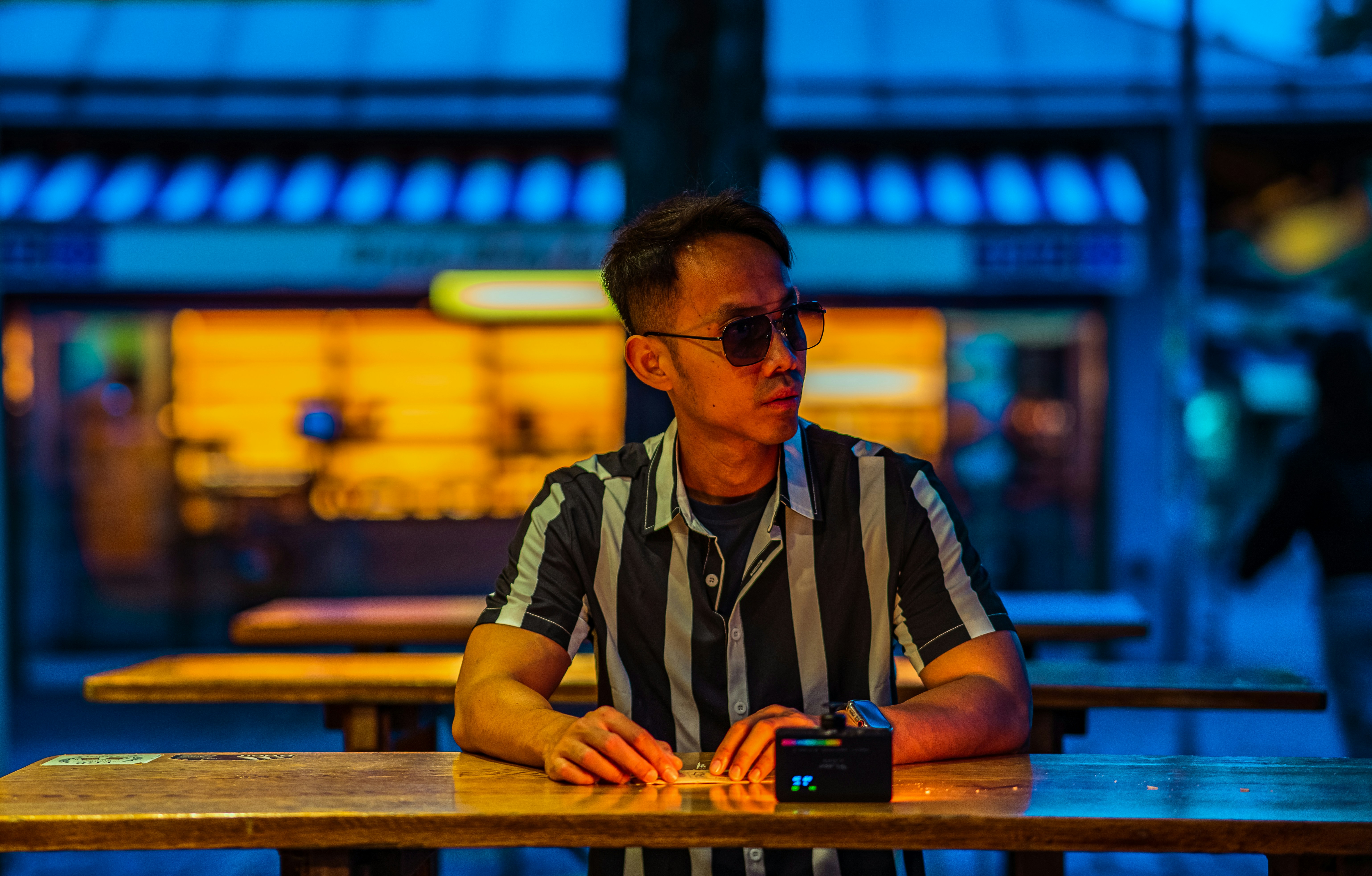A man in sunglasses sits at a wooden table, illuminated by warm lights in a bustling urban setting, with a camera placed in front of him.