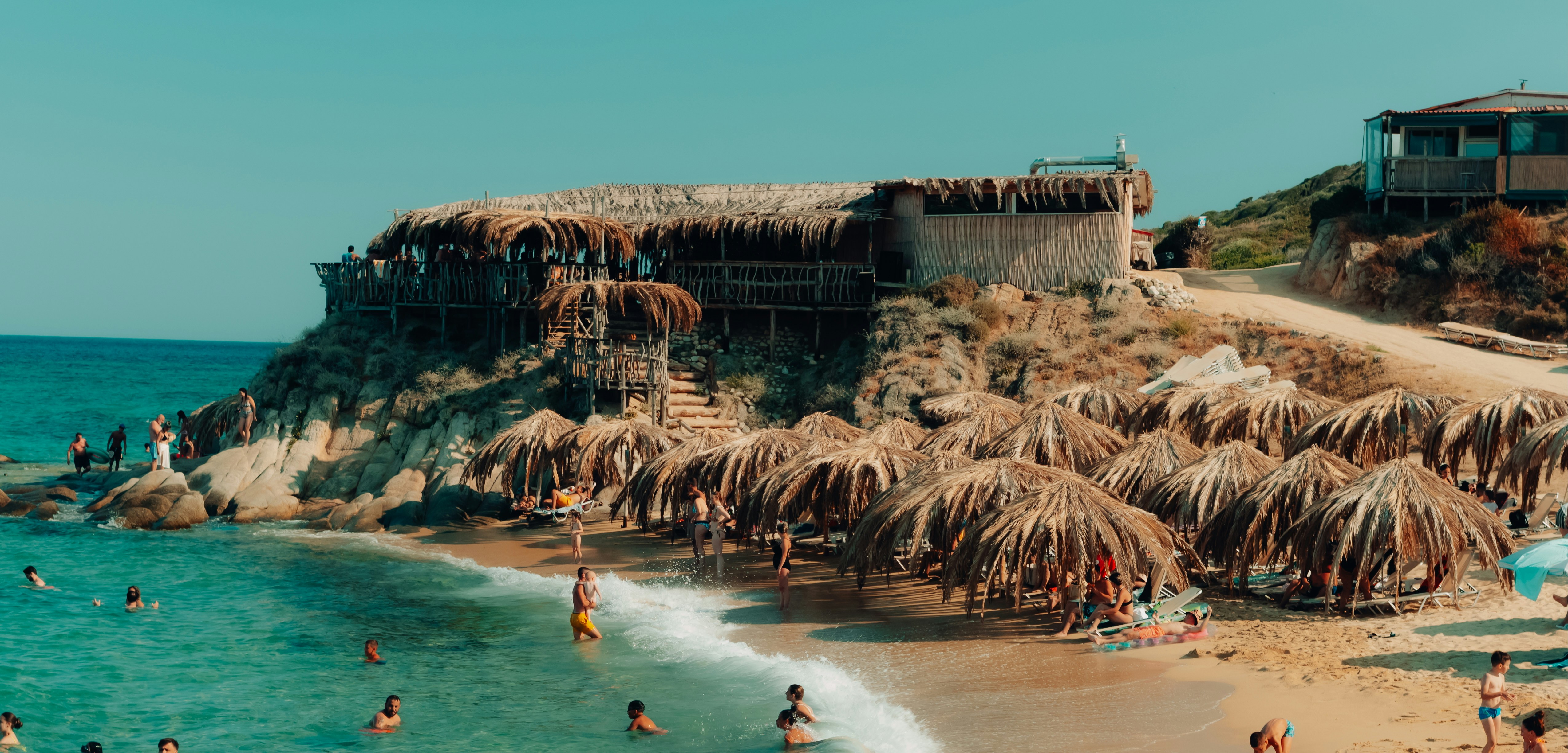 Beachgoers enjoying a sunny day under thatched umbrellas near a rustic wooden structure by the shore.