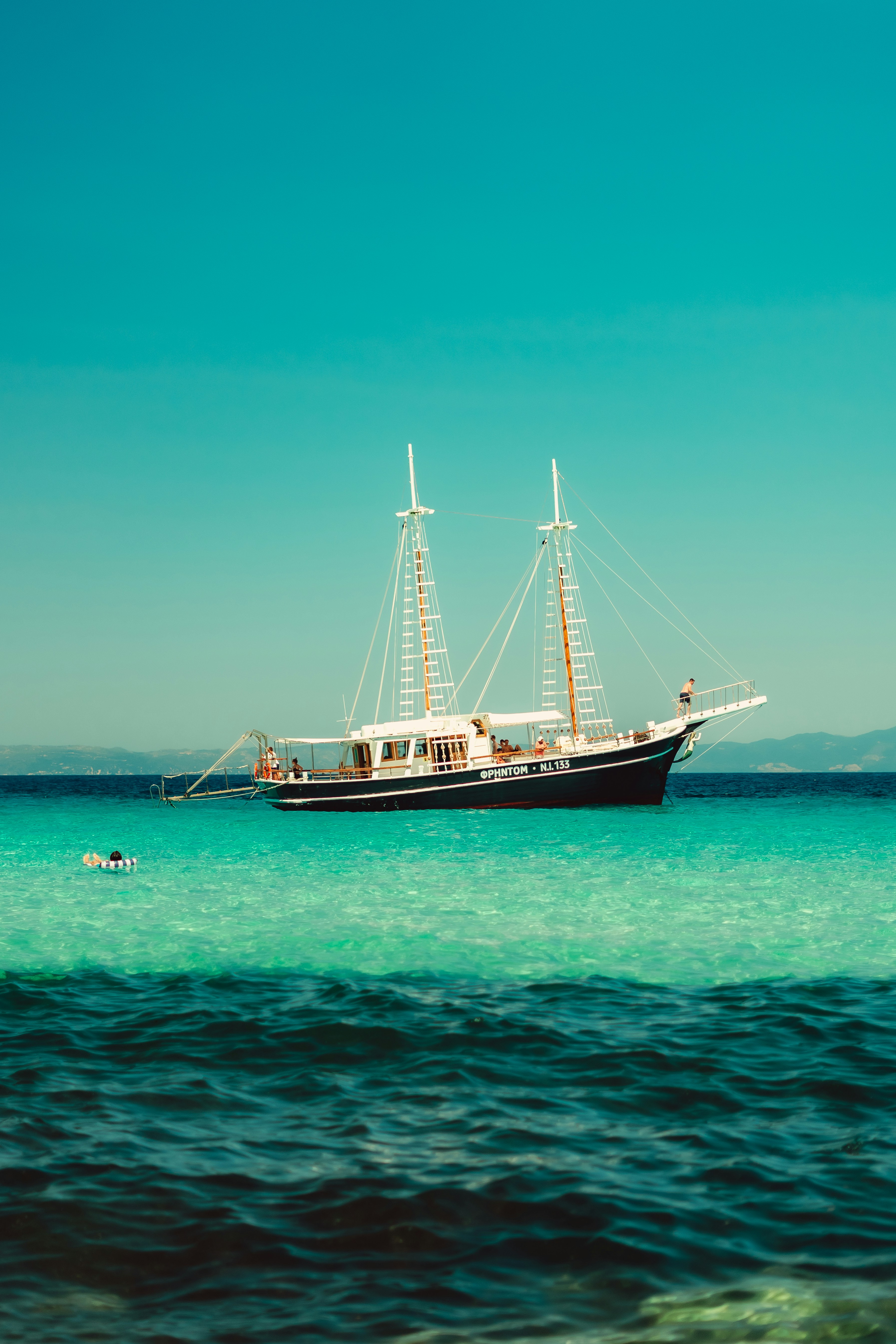 A majestic sailing ship anchored in tranquil turquoise waters, with swimmers enjoying the serene surroundings. The clear sky enhances the idyllic scene.