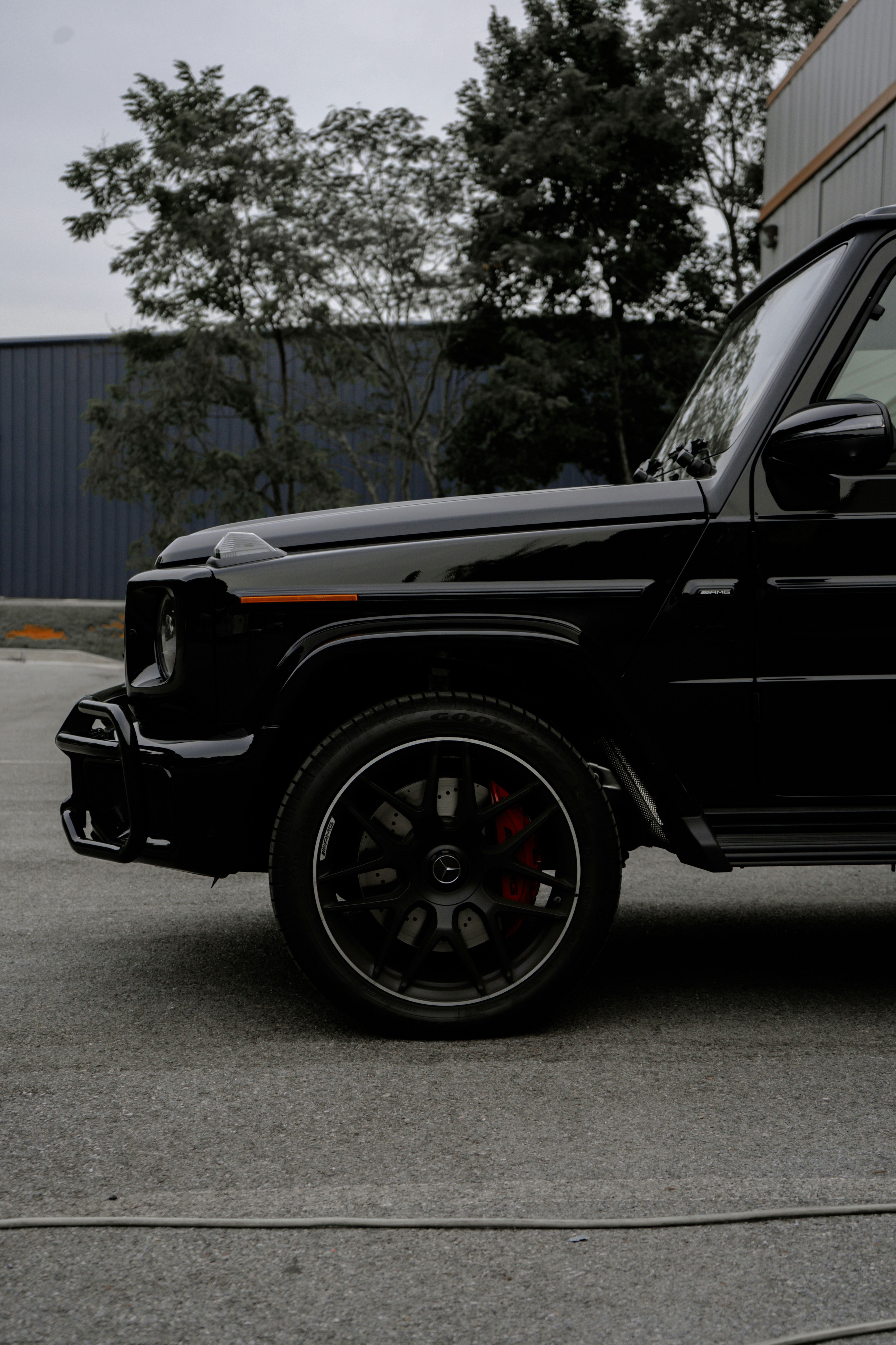 Side view of a sleek black G-Class vehicle parked on a textured surface, framed by greenery and industrial backdrop.