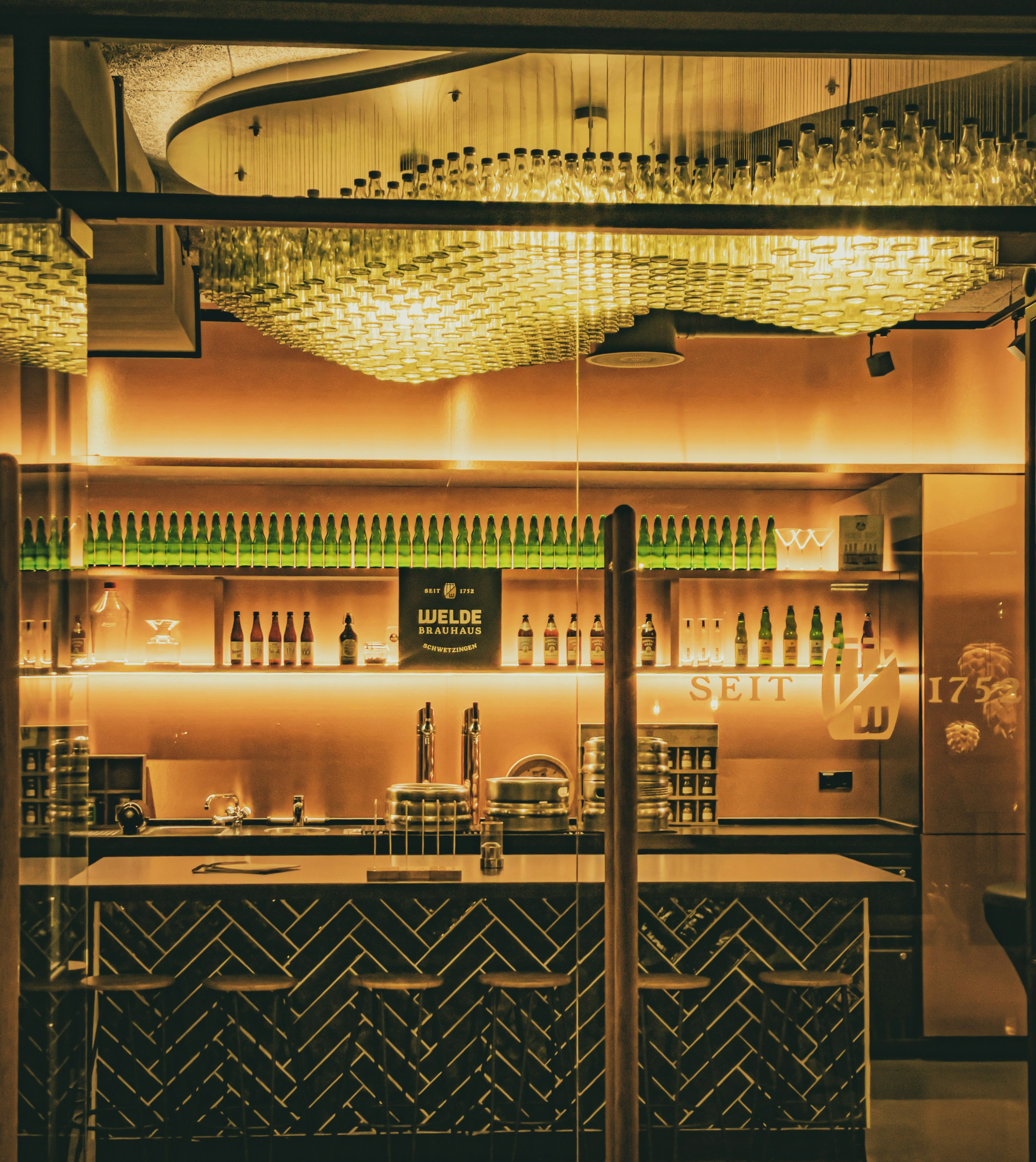 Stylish bar interior showcasing a vibrant array of bottles illuminated by warm lighting. The modern design features geometric patterns and a striking overhead installation.