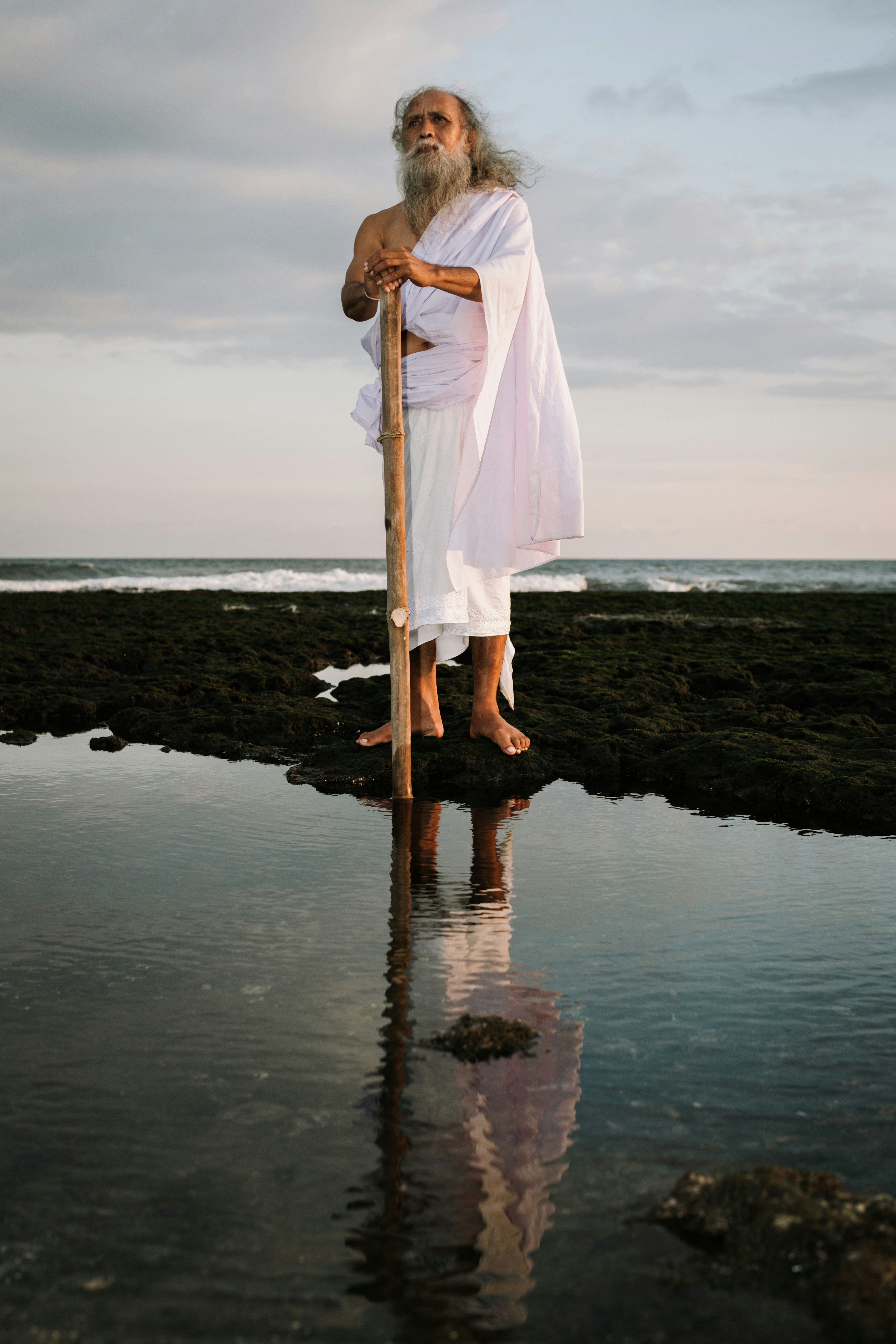 Elderly man in white robes with walking stick by ocean.