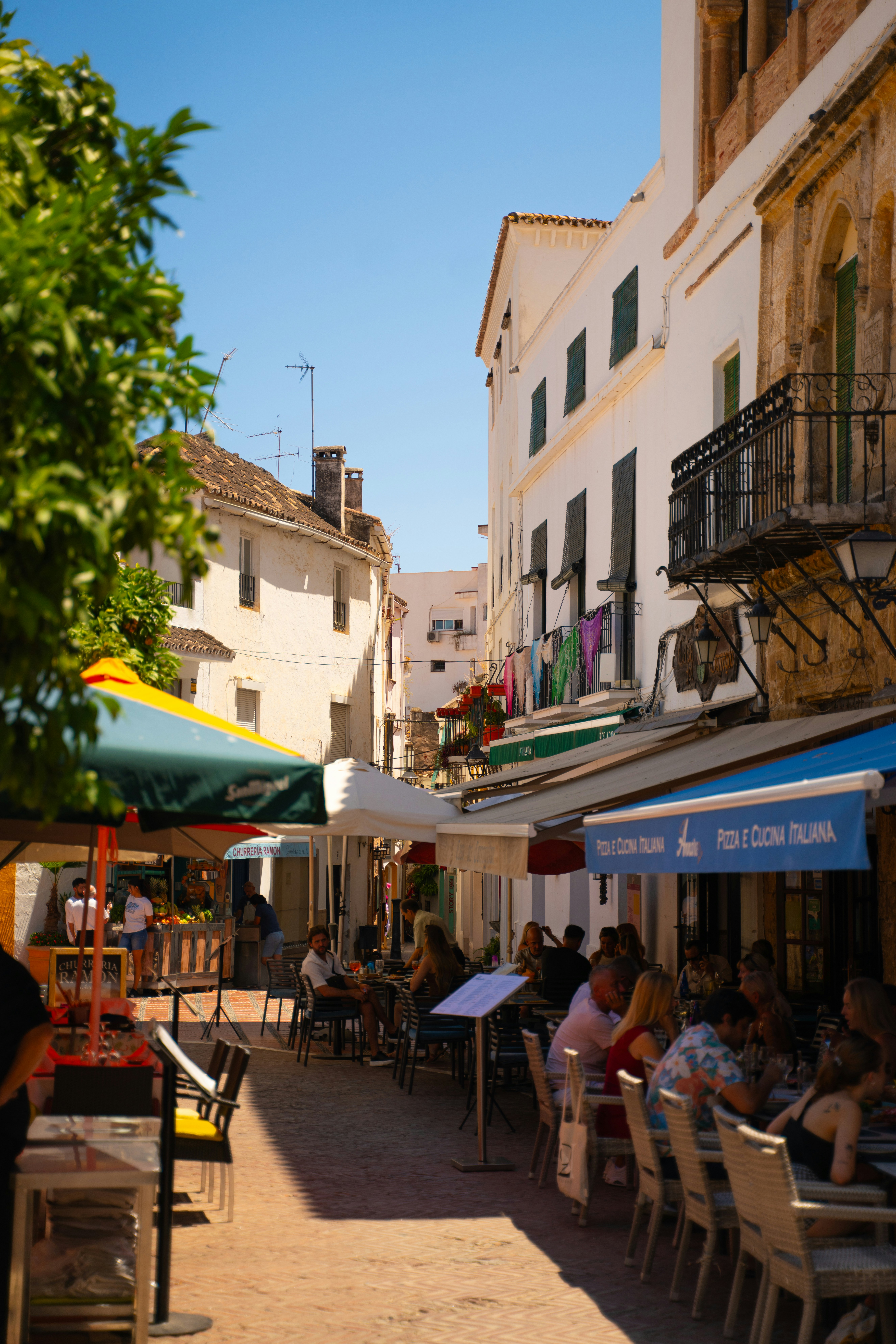 Charming alleyway lined with outdoor dining, featuring vibrant umbrellas and quaint architecture. Patrons enjoy leisurely meals under the warm sun.