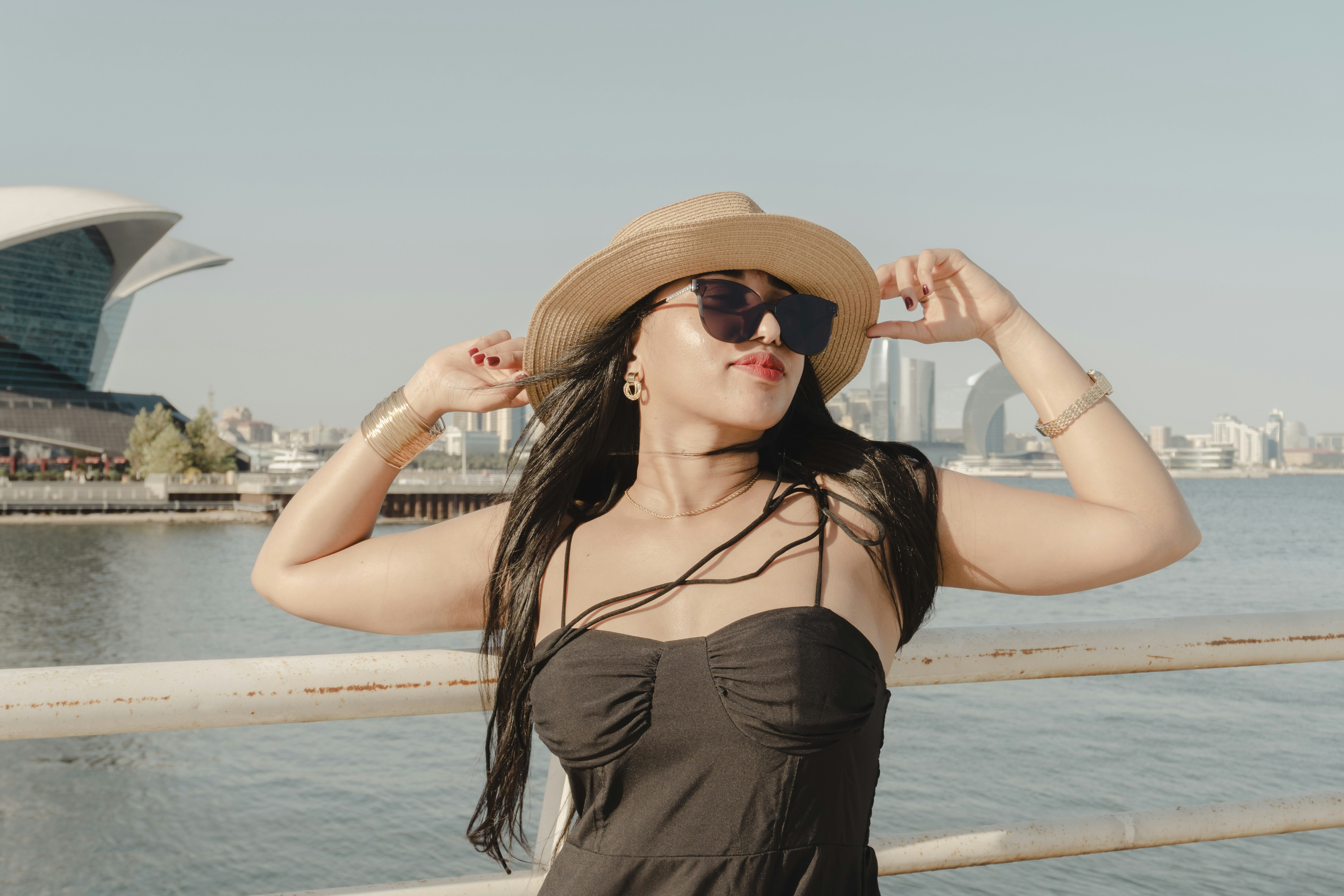 Woman in a black dress and sunhat poses confidently against a waterfront backdrop, showcasing urban architecture. The scene captures a moment of relaxation and style.