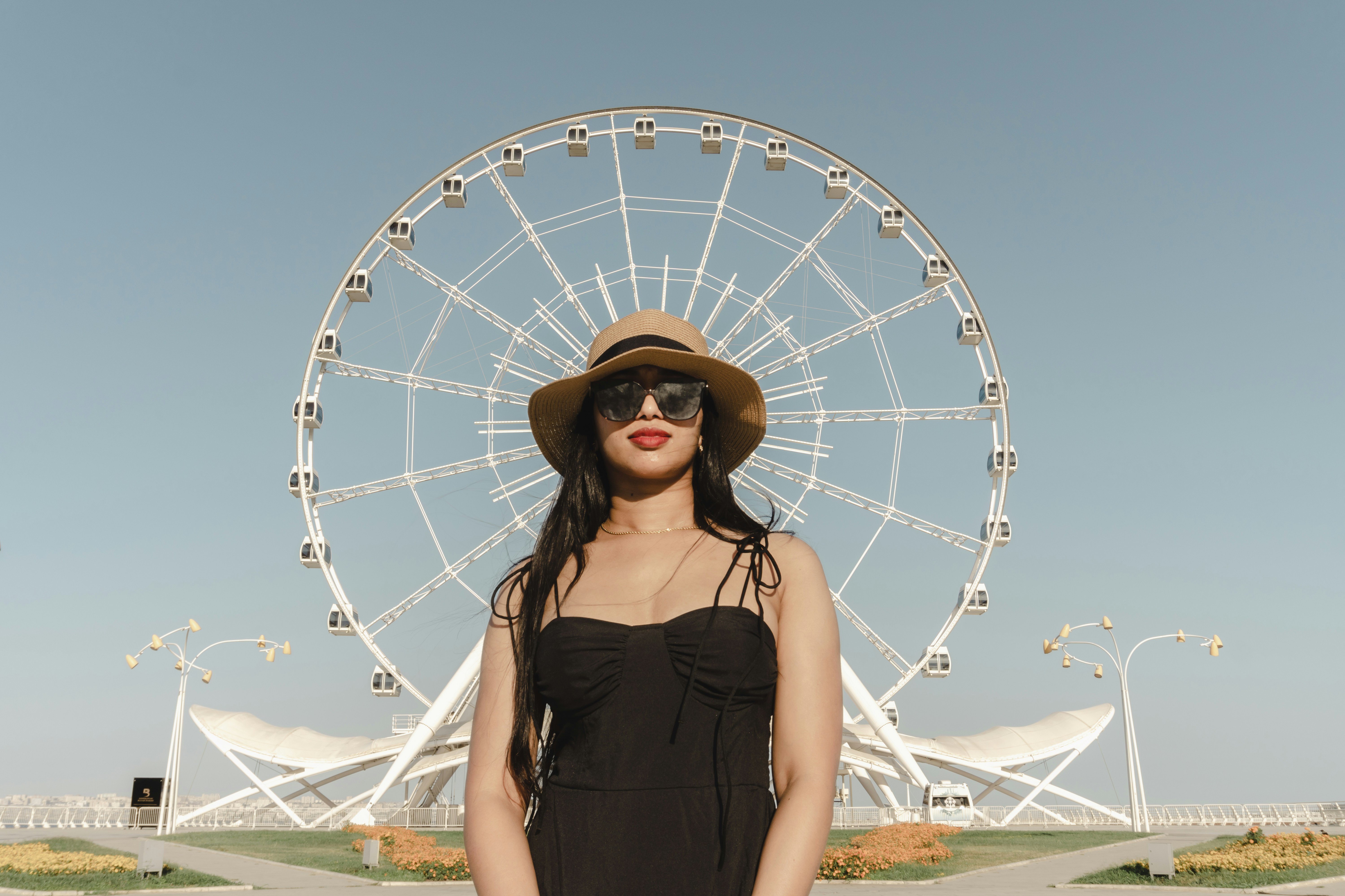 Young and beautiful female traveler wearing a stylish dress and summer hat enjoys evening ambience at the Baku Boulevard | Woman in hat and sunglasses in front of ferris wheel