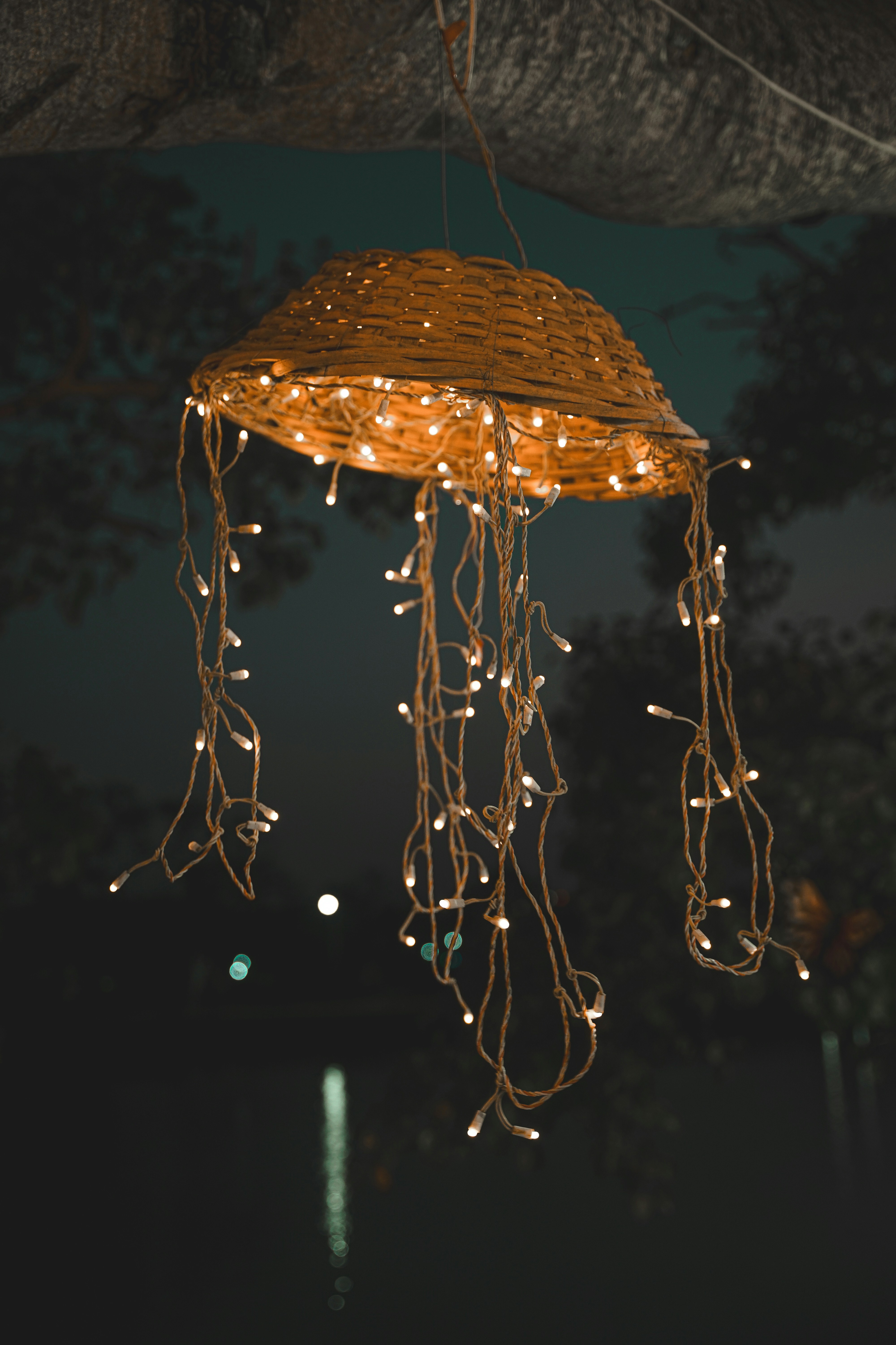 A woven basket-shaped light fixture adorned with fairy lights, suspended from a tree branch against a dark backdrop. The soft glow creates an enchanting atmosphere.