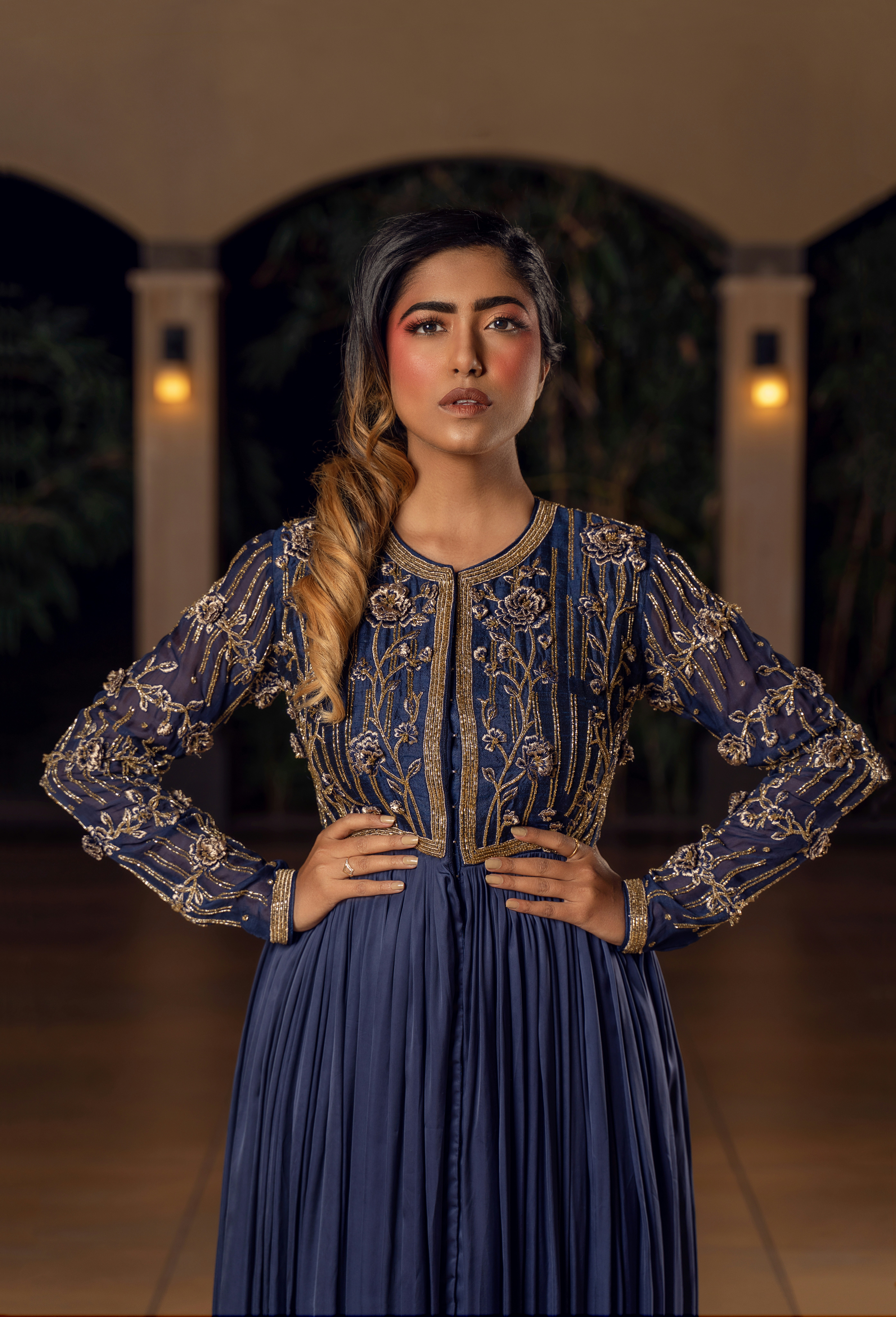 A poised individual in an intricately embroidered blue dress stands confidently, showcasing the artistry of traditional attire against a softly lit backdrop.