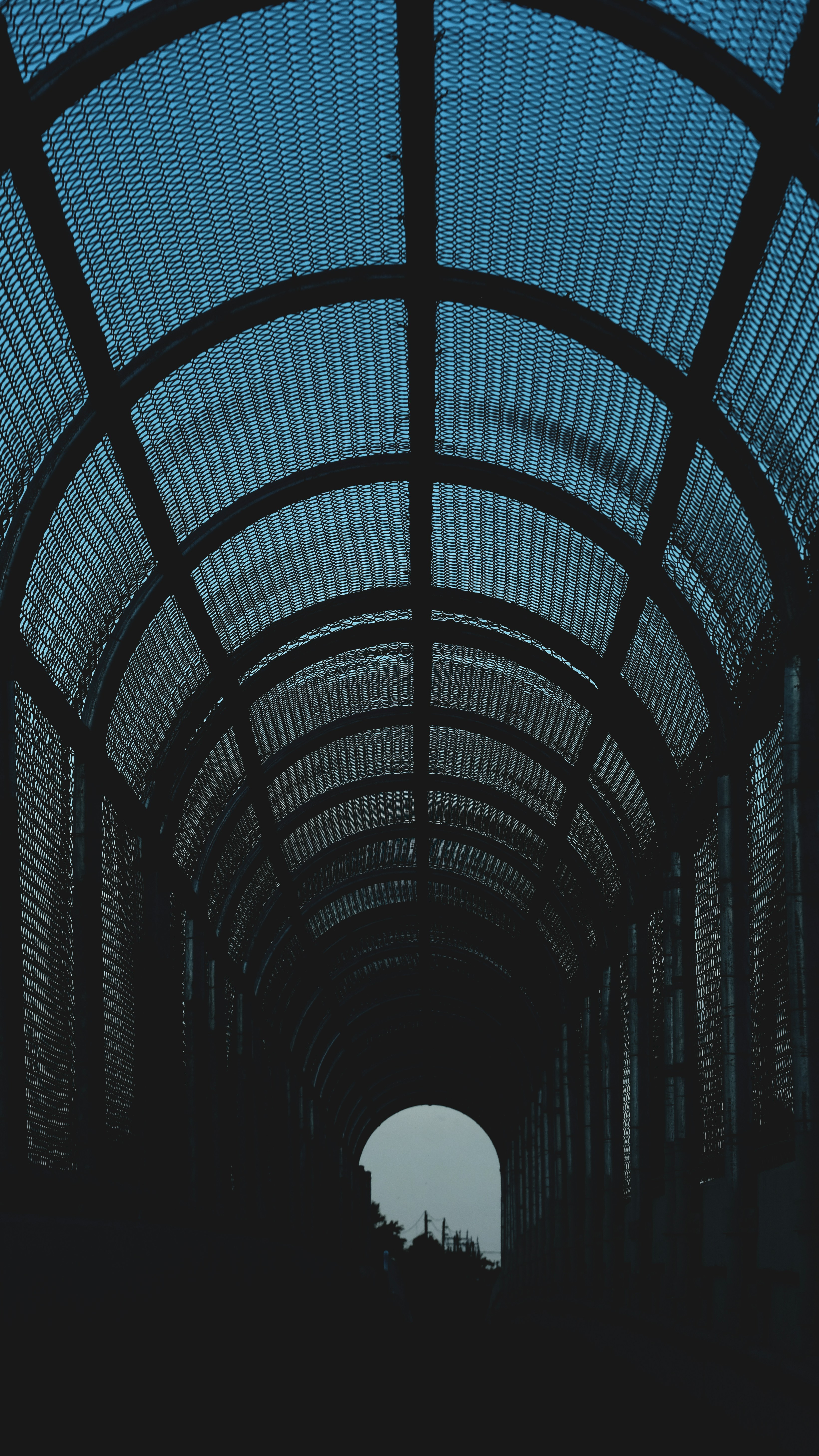 Image of a metal mesh tunnel on a bridge, with an arched grid ceiling leading to a bright exit. | Dark arched tunnel with blue ceiling and mesh