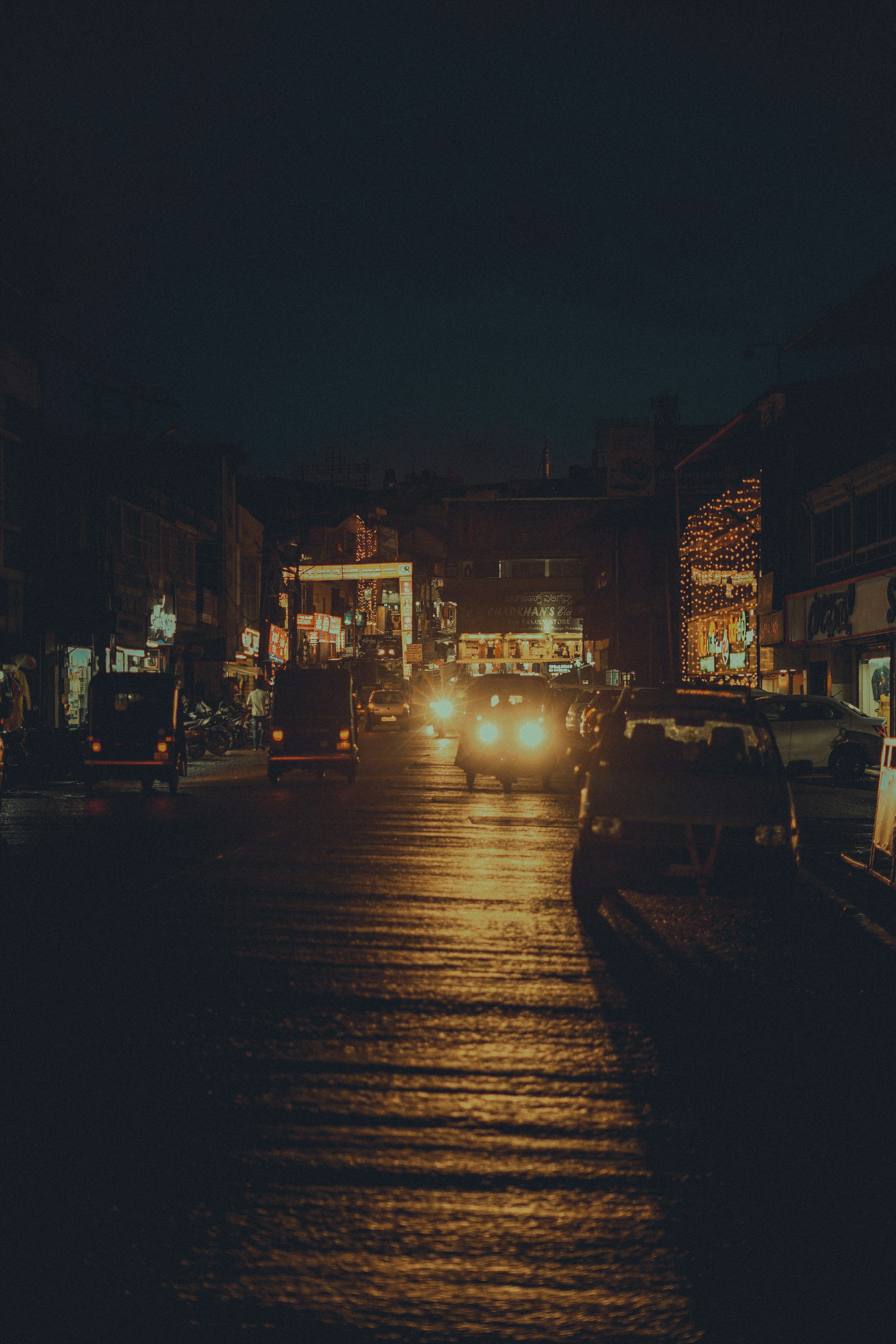 busy city street glowing with headlights and shop lights at night | Cars driving on wet street at night