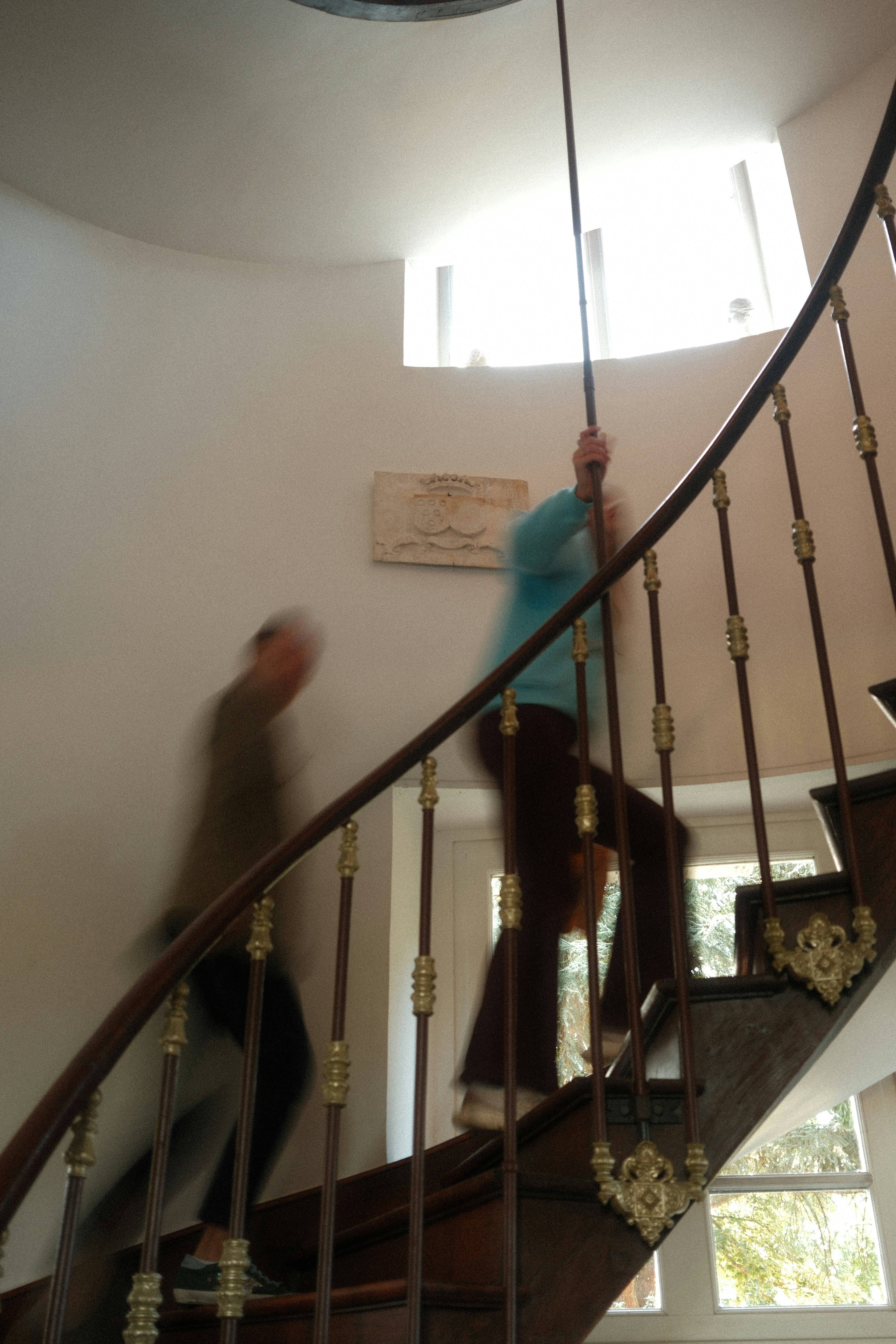 Two figures gracefully navigate a spiral staircase, their movements captured in a blur against a soft-lit backdrop.