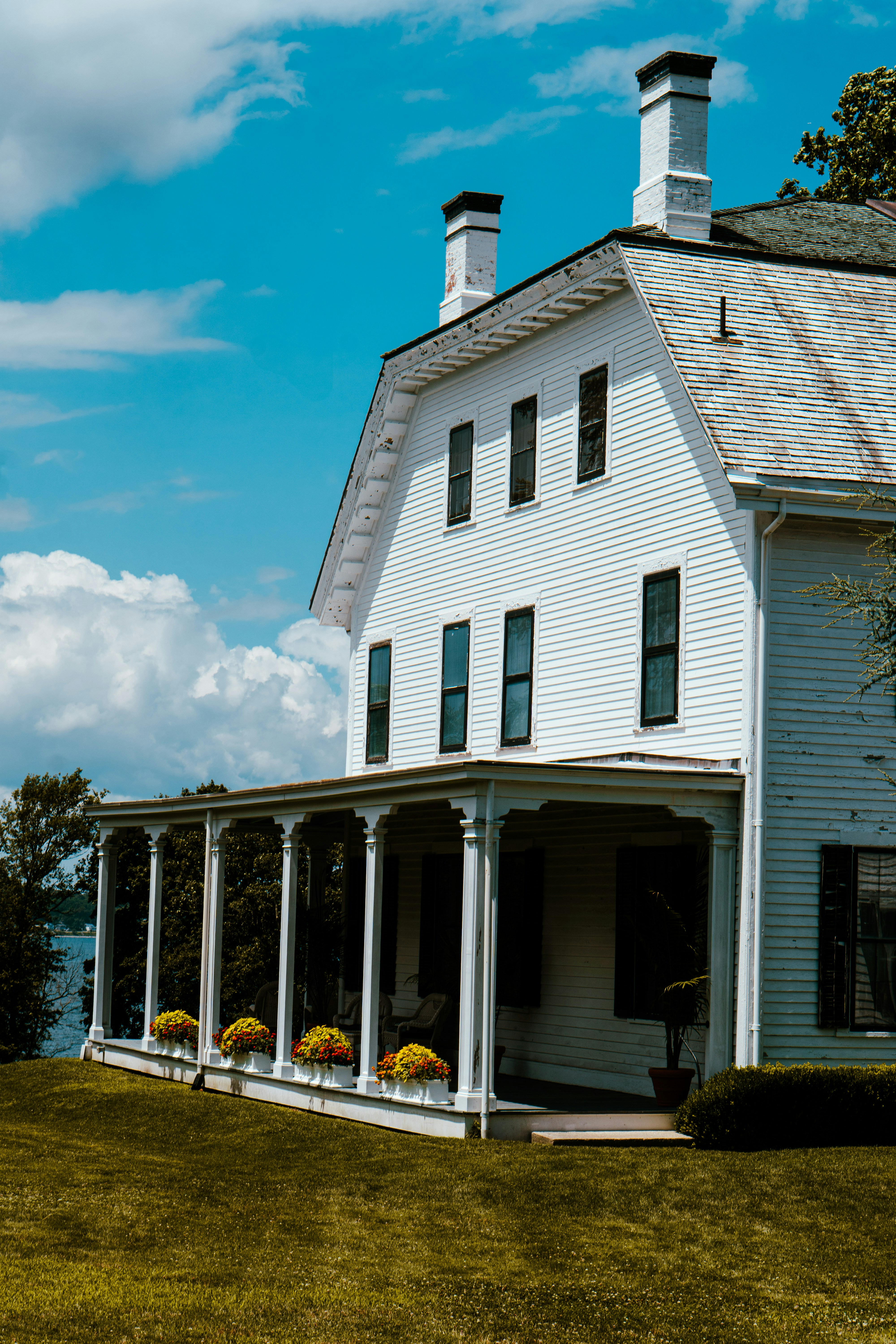 Historic white house with a wraparound porch adorned with vibrant flowers, set against a backdrop of blue skies and fluffy clouds.