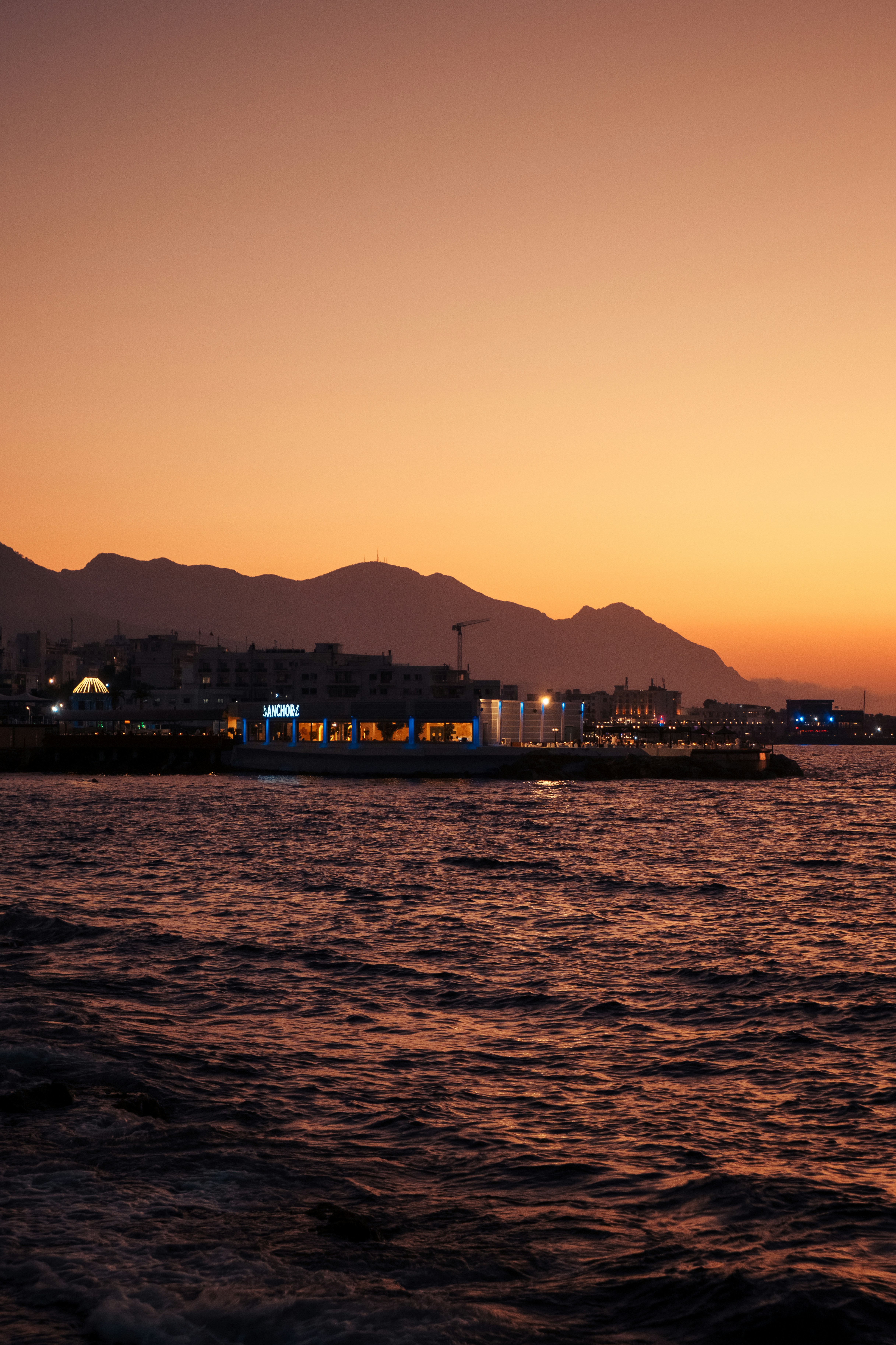 A serene coastal scene at twilight, showcasing illuminated buildings along the waterfront against a backdrop of distant mountains. The gentle waves reflect the warm hues of the sunset.