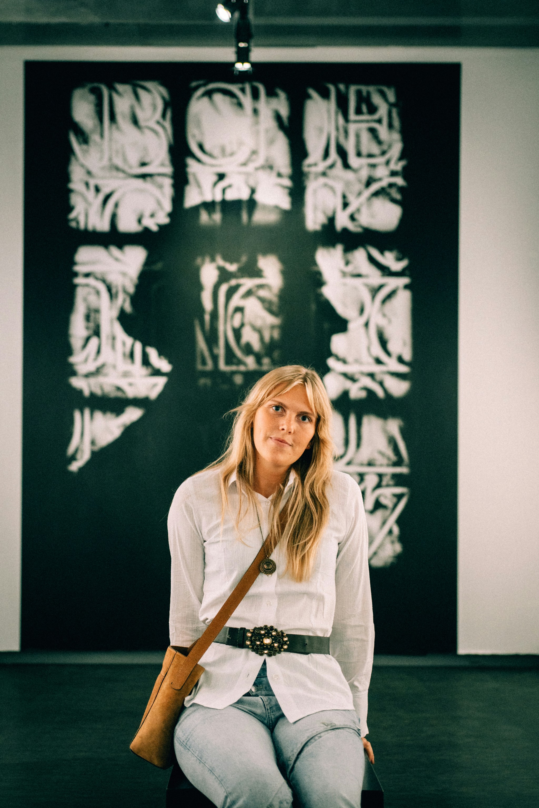 A woman seated in an art gallery, gazing thoughtfully at a large abstract mural behind her. Her casual attire contrasts with the bold artwork.