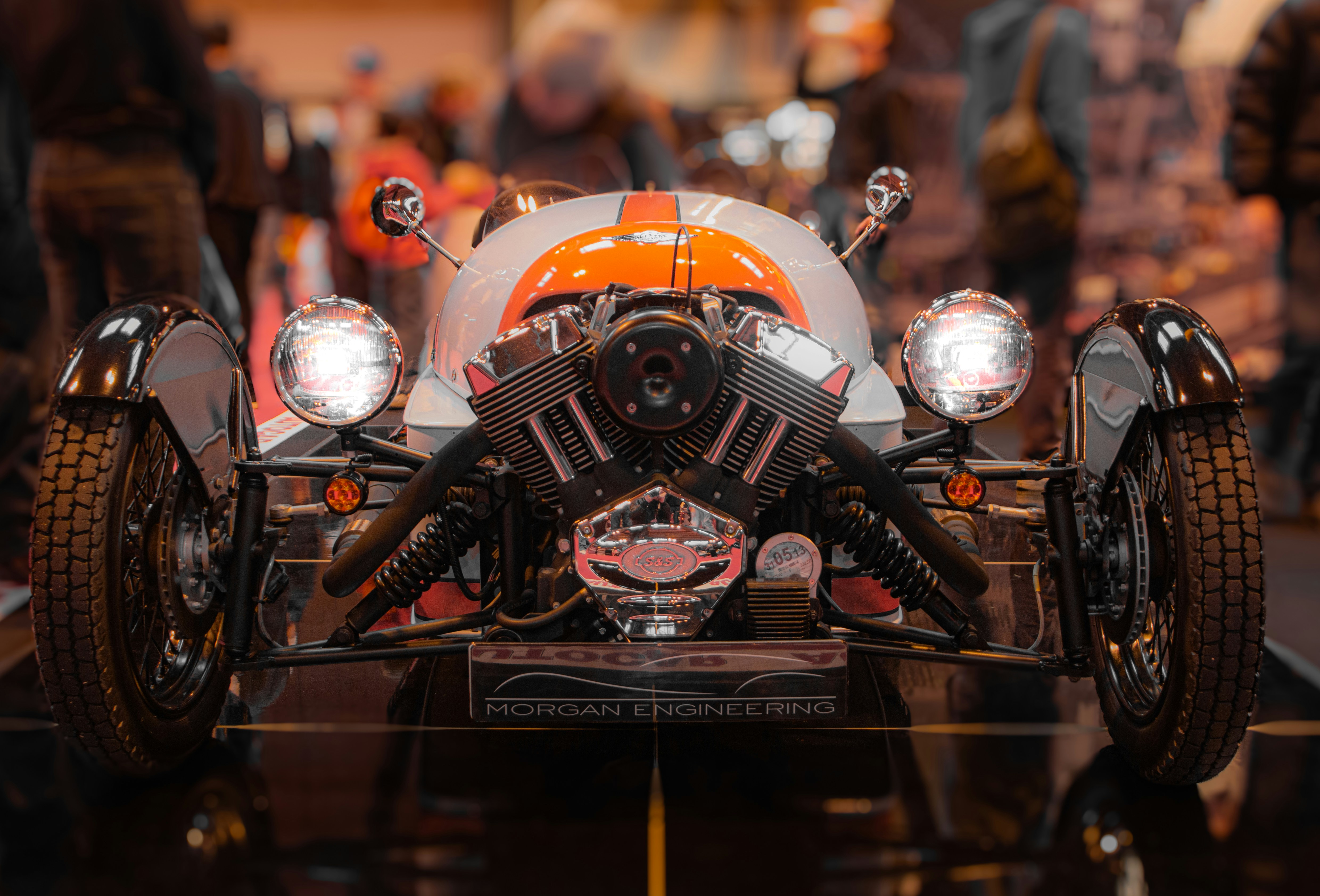 Front view of a Morgan three-wheeler, showcasing its exposed engine and unique retro design at a car show. | Front view of a three-wheeled vintage car