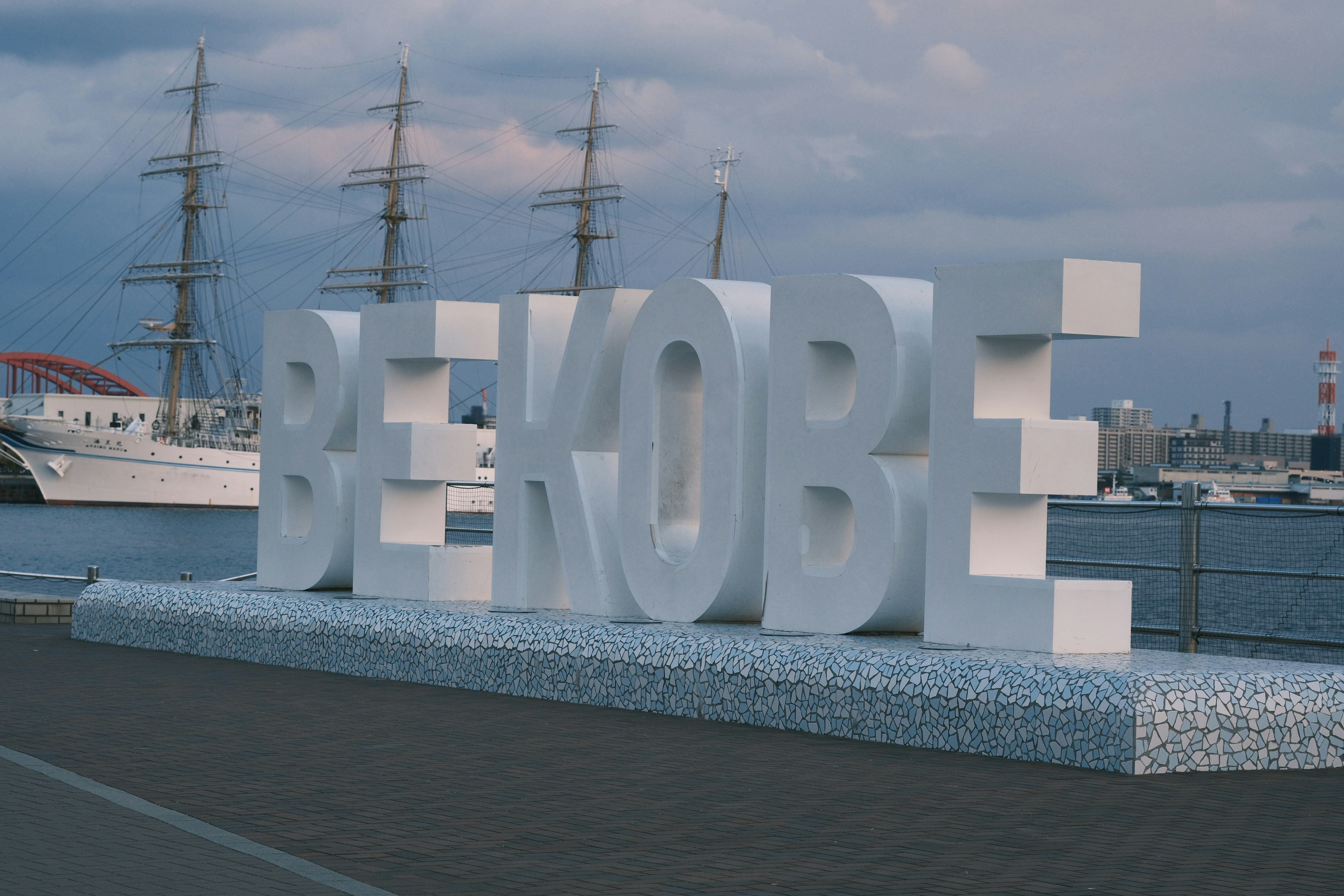 Bold white letters spelling 'BE KOBE' stand prominently along the waterfront, with historical ships and a cloudy sky in the background.