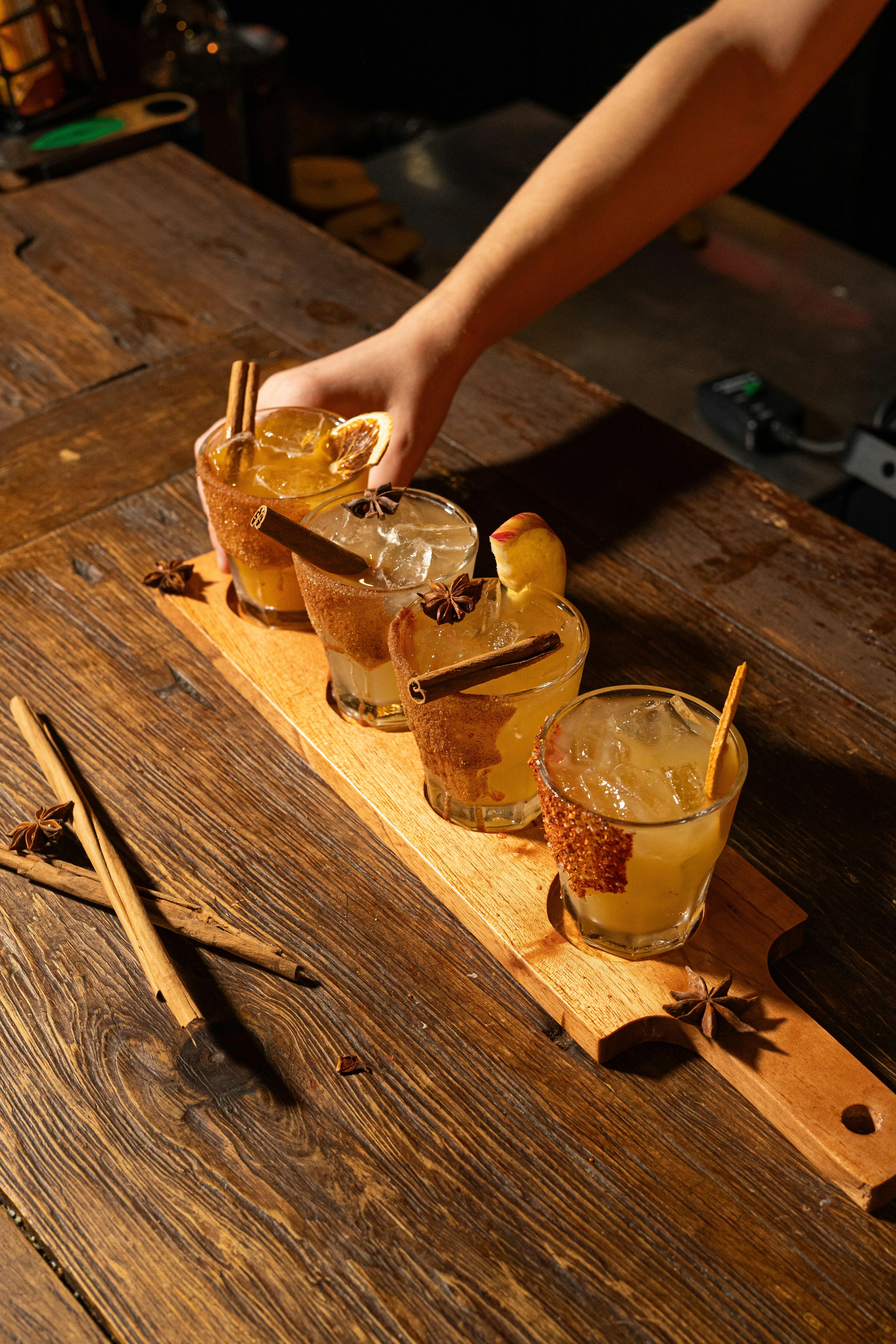 Four elegantly garnished cocktails arranged on a wooden platter, with a hand reaching to serve. The scene highlights the artistry of mixology.