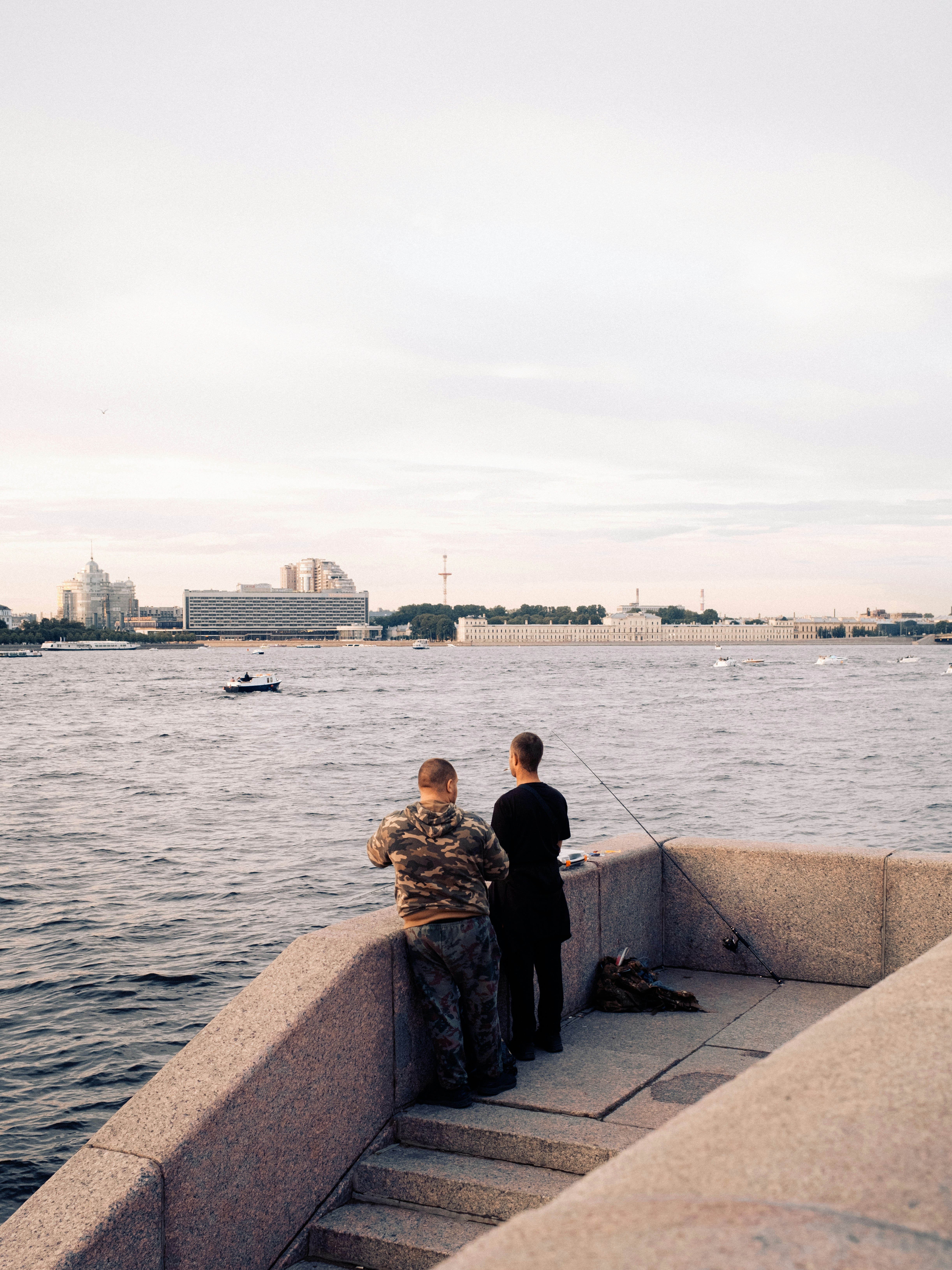 Two fishermen enjoy a quiet moment on a stone pier, casting their lines into the tranquil waters as the city skyline looms in the background.
