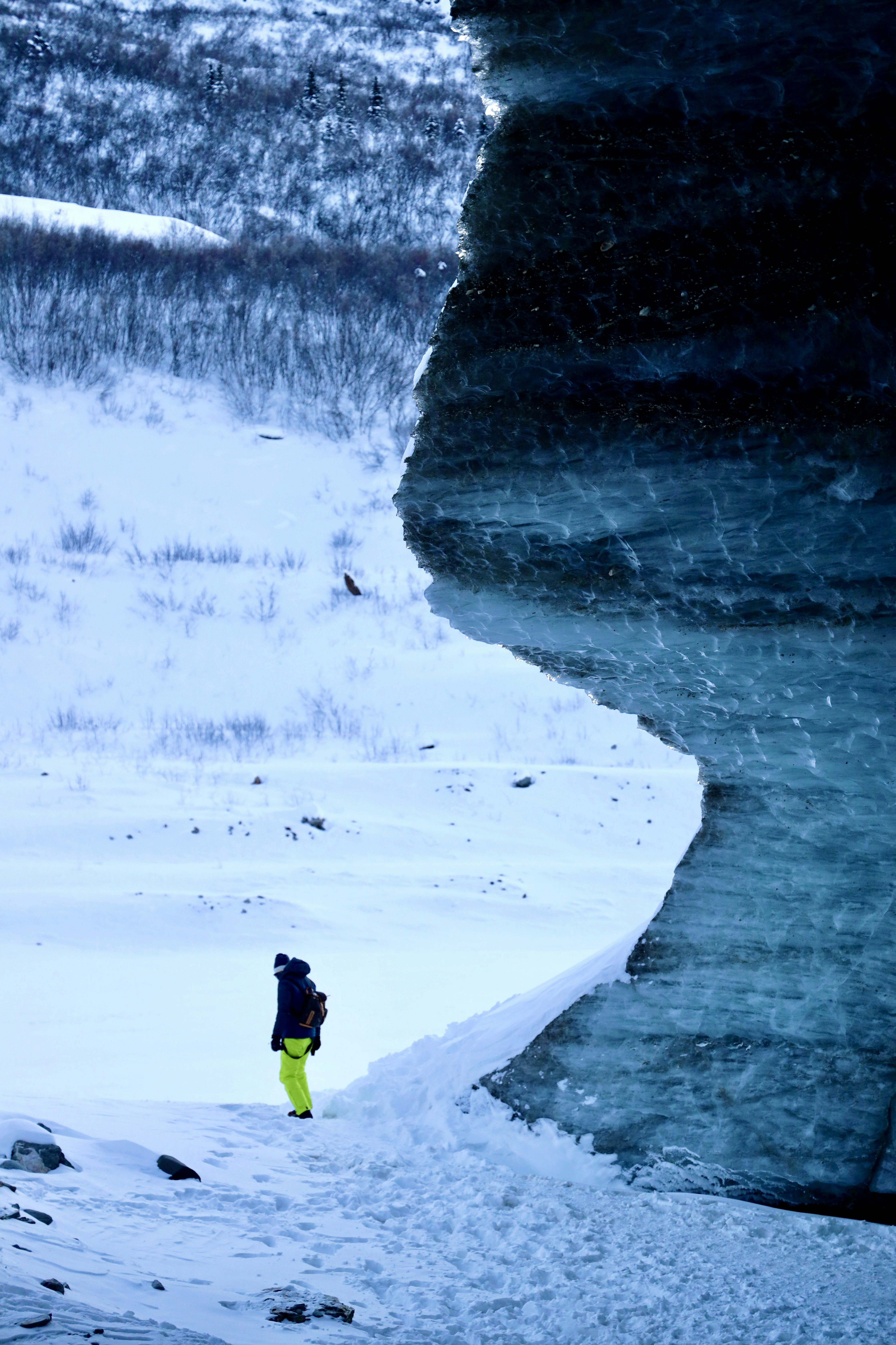 A lone explorer dwarfed by the towering walls of an Alaskan ice cave — a meeting of scale, silence, and raw adventure | Person standing by a large blue ice formation in snow.