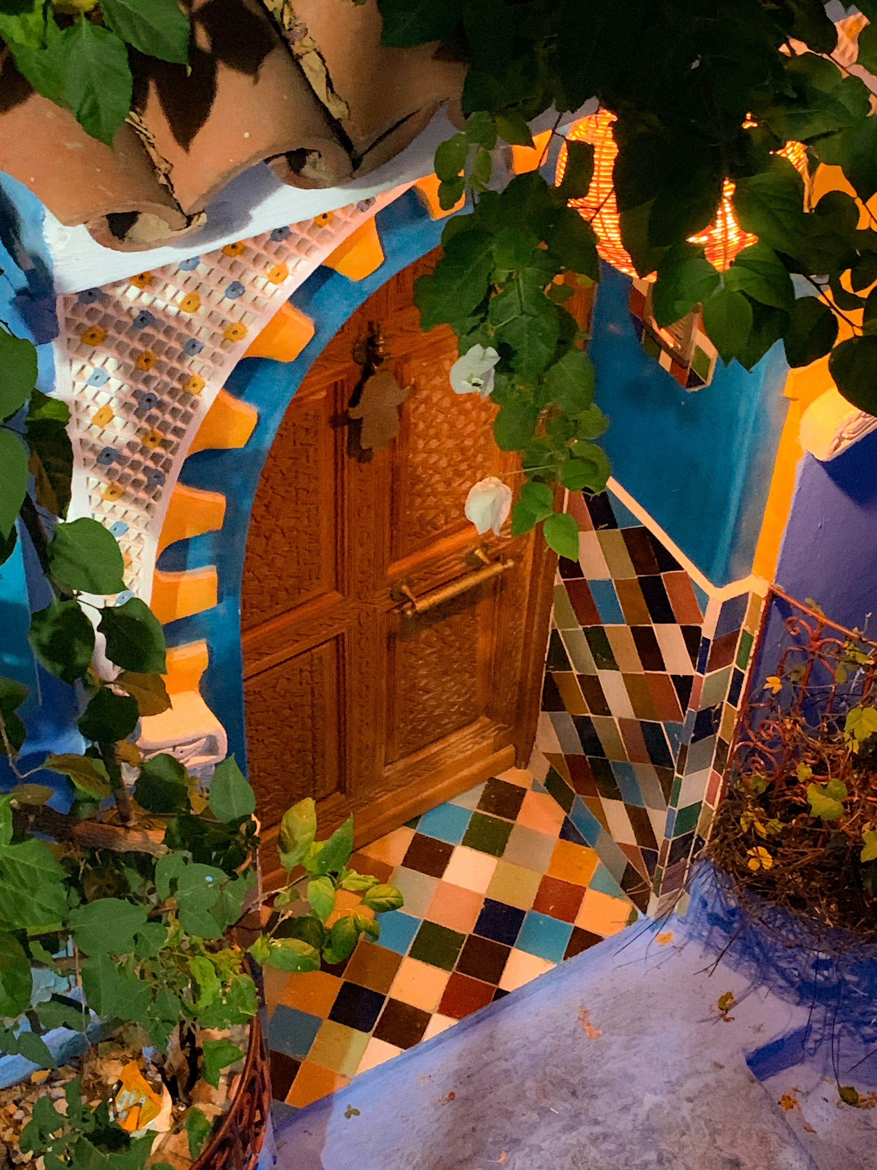Wandering the streets of Chefchaouen is like stepping into a dream painted in a thousand shades of blue. From deep cobalt to soft sky, the radiant hues wash over every wall, step, and doorway, creating a serene and cool labyrinth that stands in breathtaking contrast to the warm, earthy tones of the surrounding Rif Mountains. The beauty is in the details: a vibrant magenta bougainvillea spilling over an azure wall, the intricate patterns of a handwoven rug hanging beside a cerulean alleyway, and the soft play of light and shadow that transforms t | Colorful tiled doorway with wooden door and plants