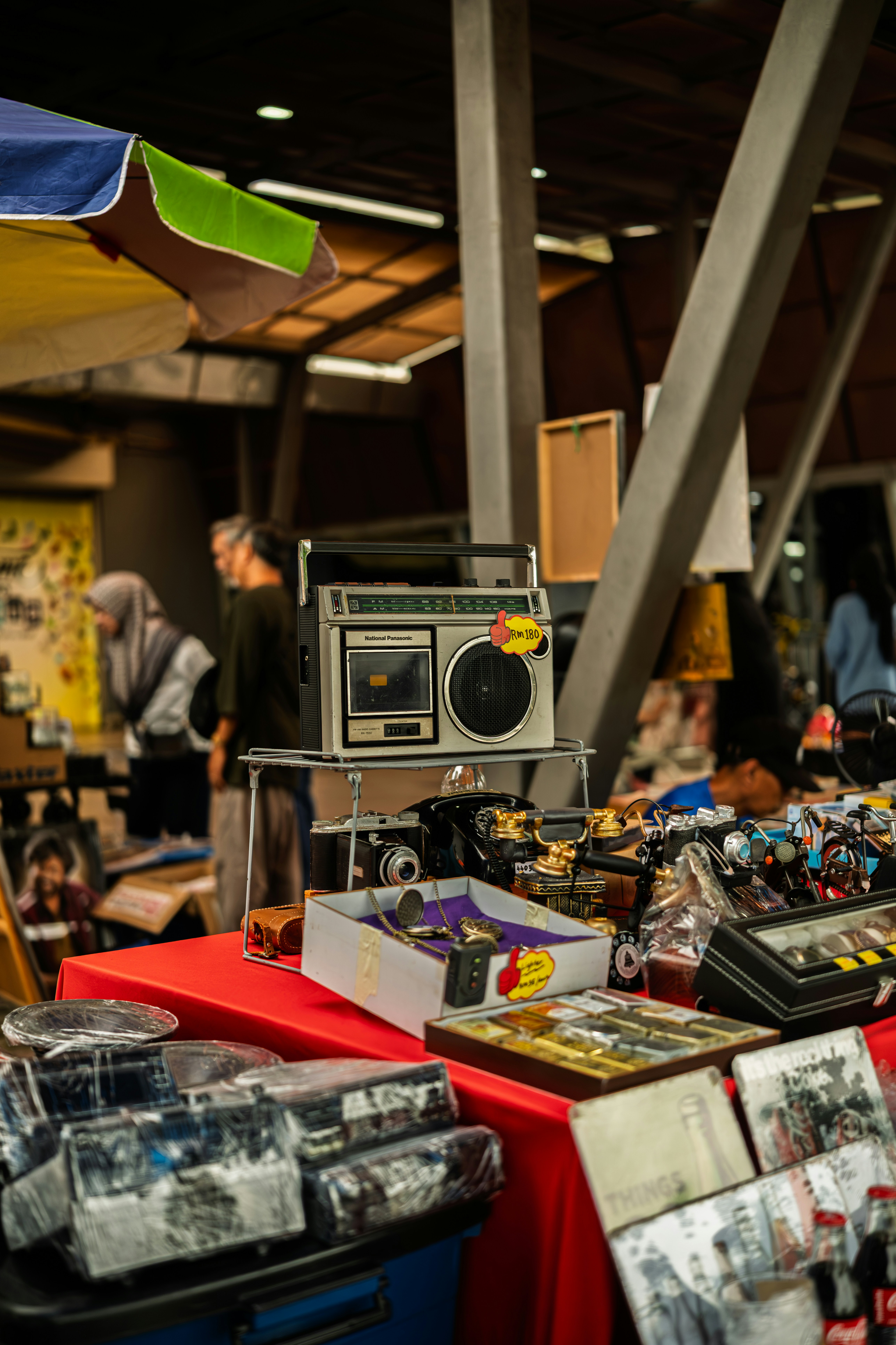 Vintage radio and assorted trinkets displayed on a vibrant red table at a bustling market. The background features shoppers browsing various stalls.