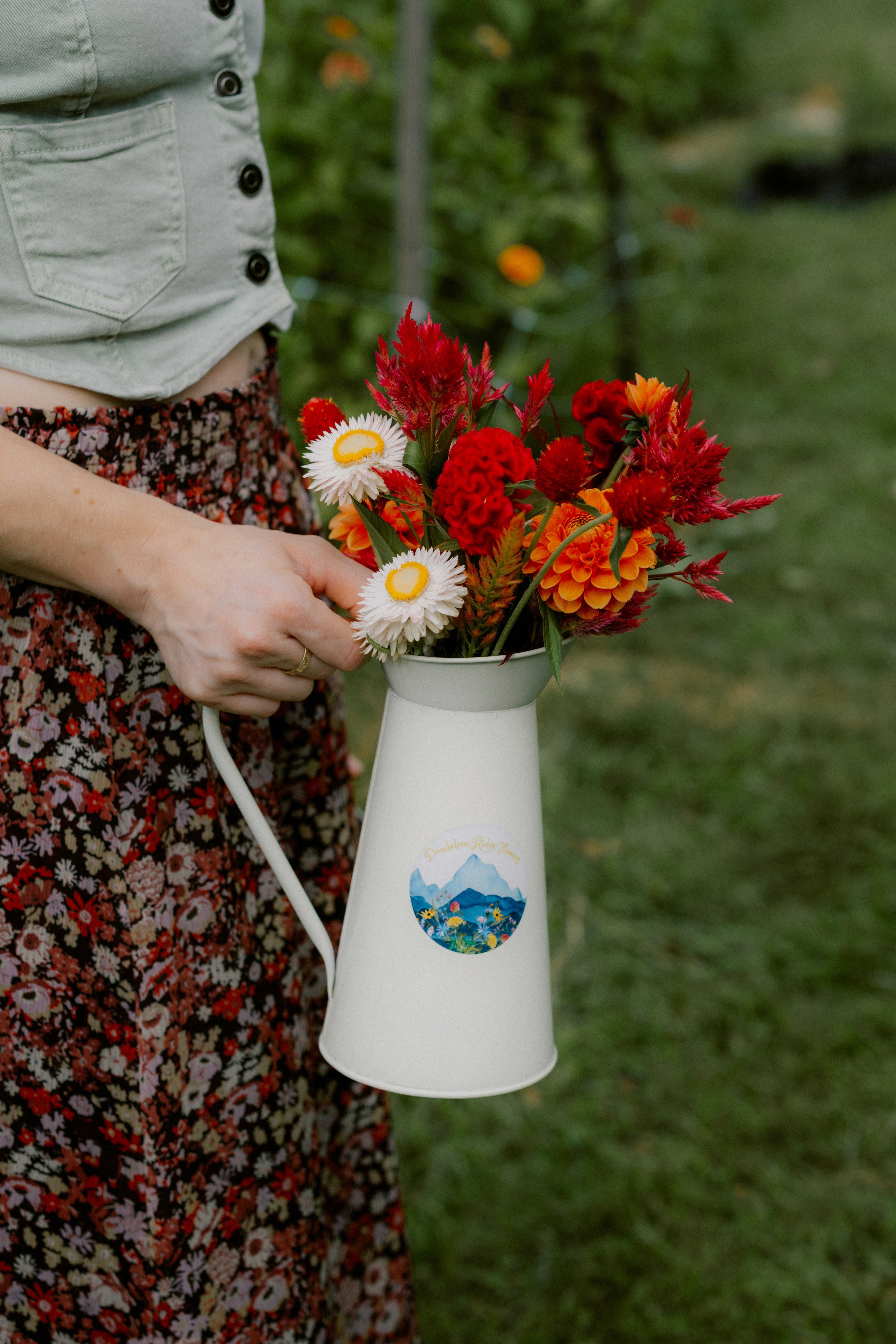 A person in a floral dress holds a white pitcher overflowing with a vivid bouquet outdoors.