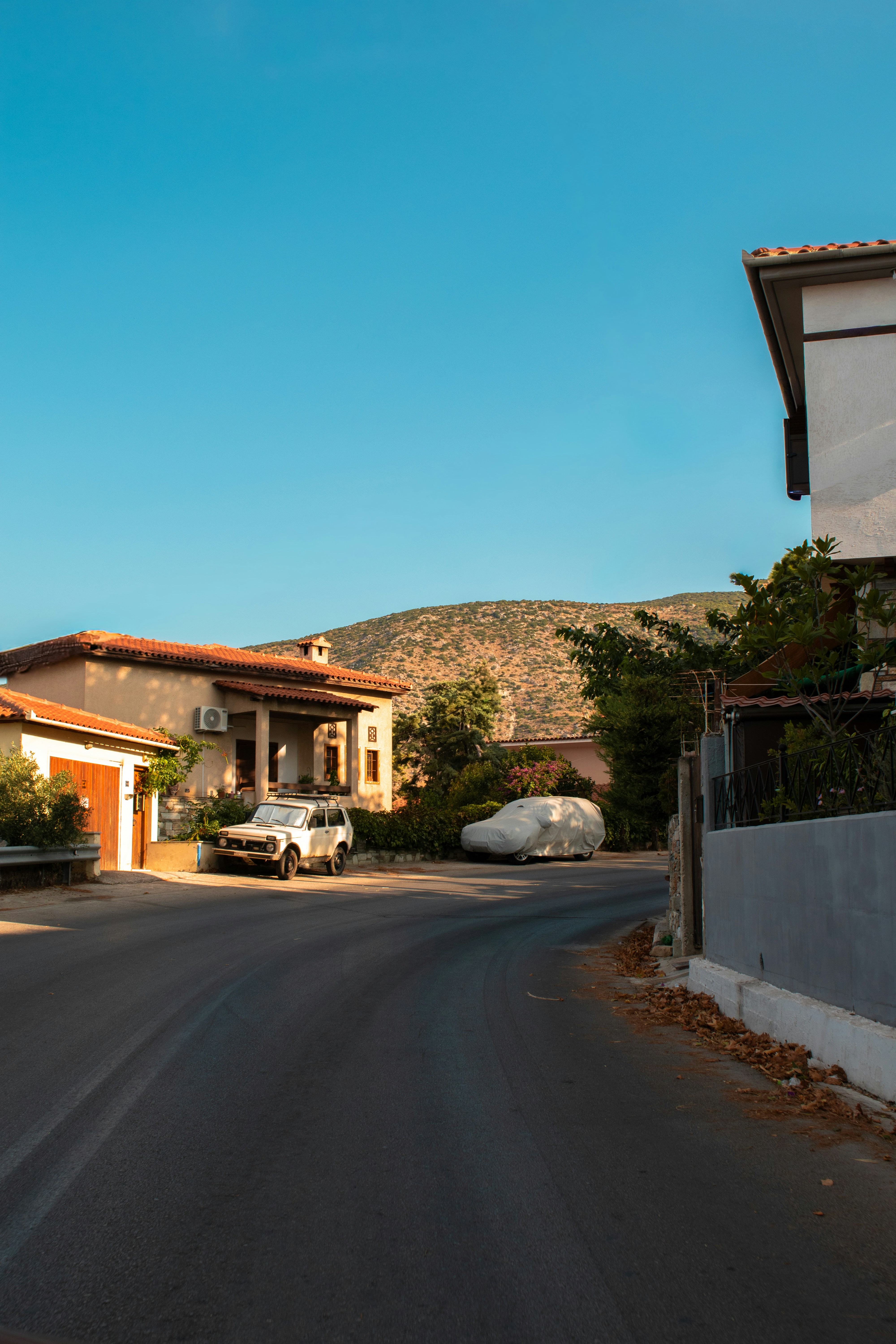 Charming village street with traditional houses and a parked vehicle, framed by rolling hills under a clear blue sky.
