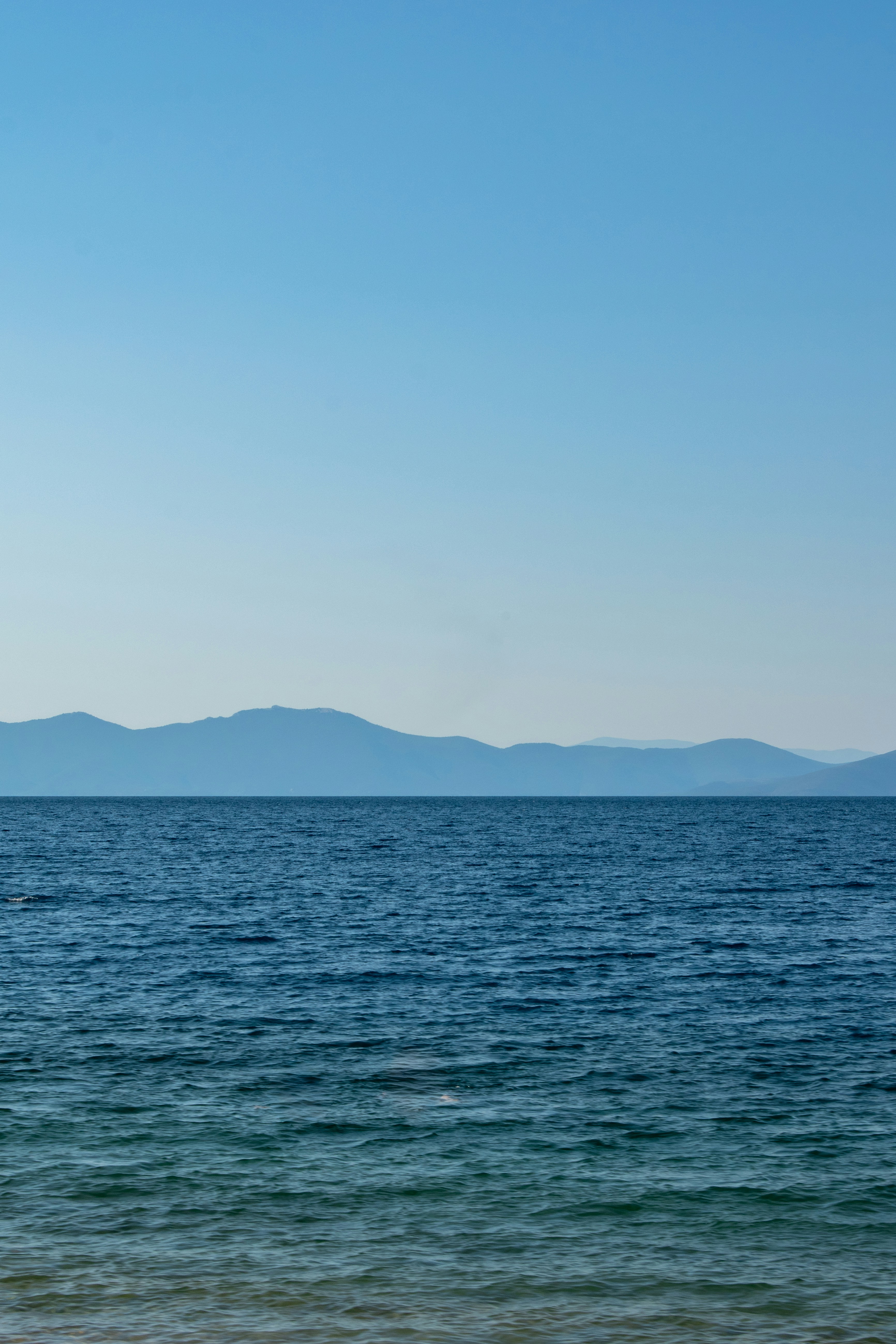 Gentle waves lapping against the shore under a clear blue sky, with distant mountains softly fading into the horizon.