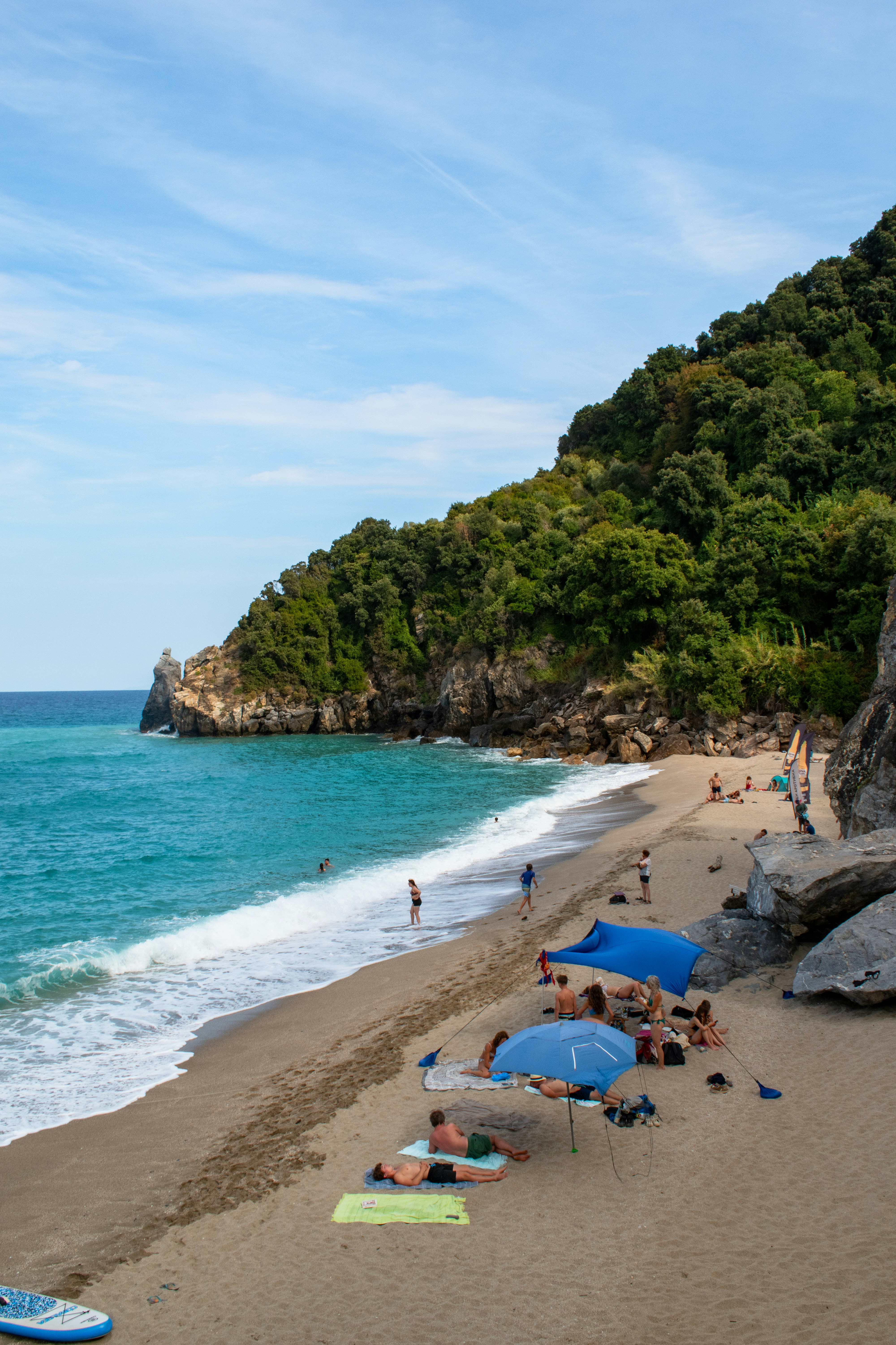 Secluded beach scene featuring sunbathers and umbrellas, framed by lush greenery and rocky cliffs. Waves gently lap at the shore.