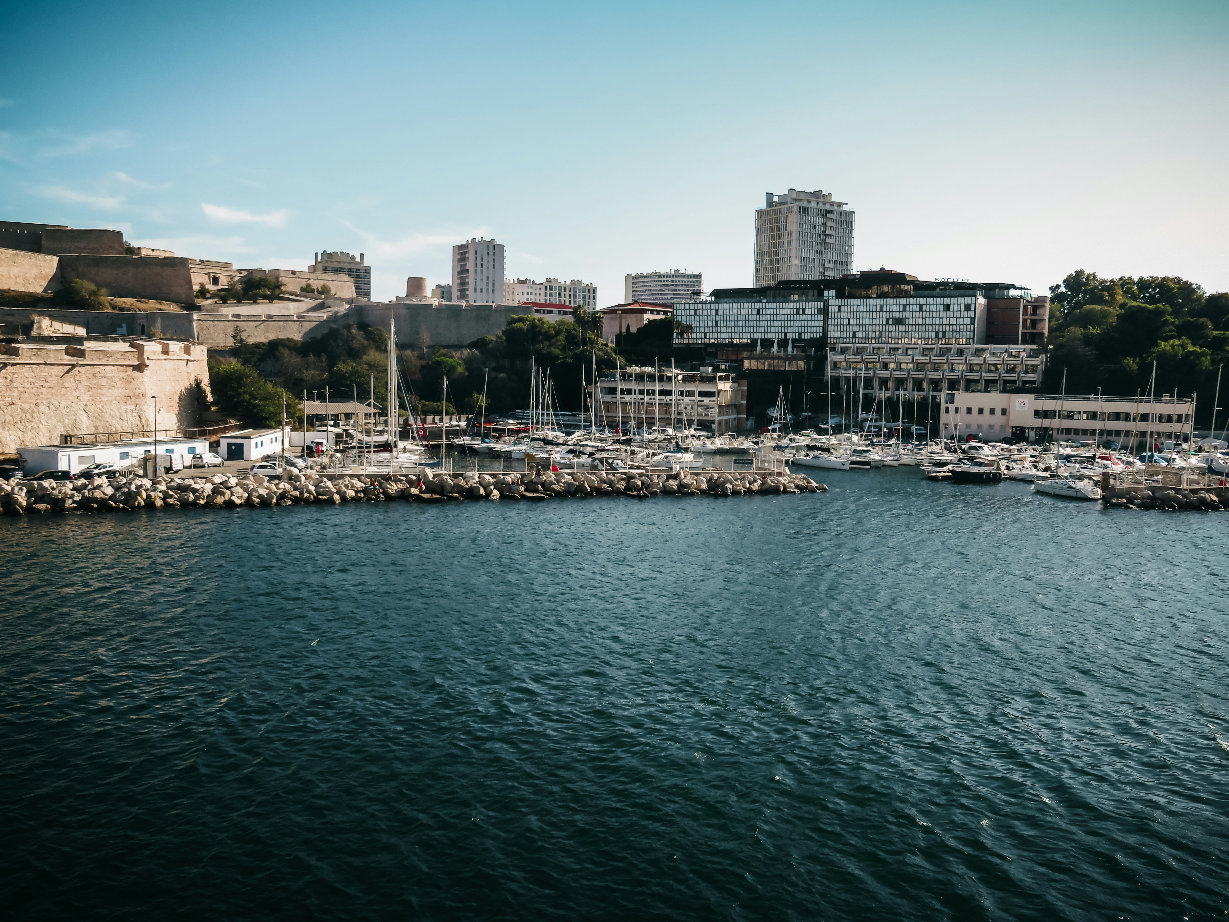 Marina filled with sailboats alongside a coastal town, framed by historical fortifications and modern buildings. 