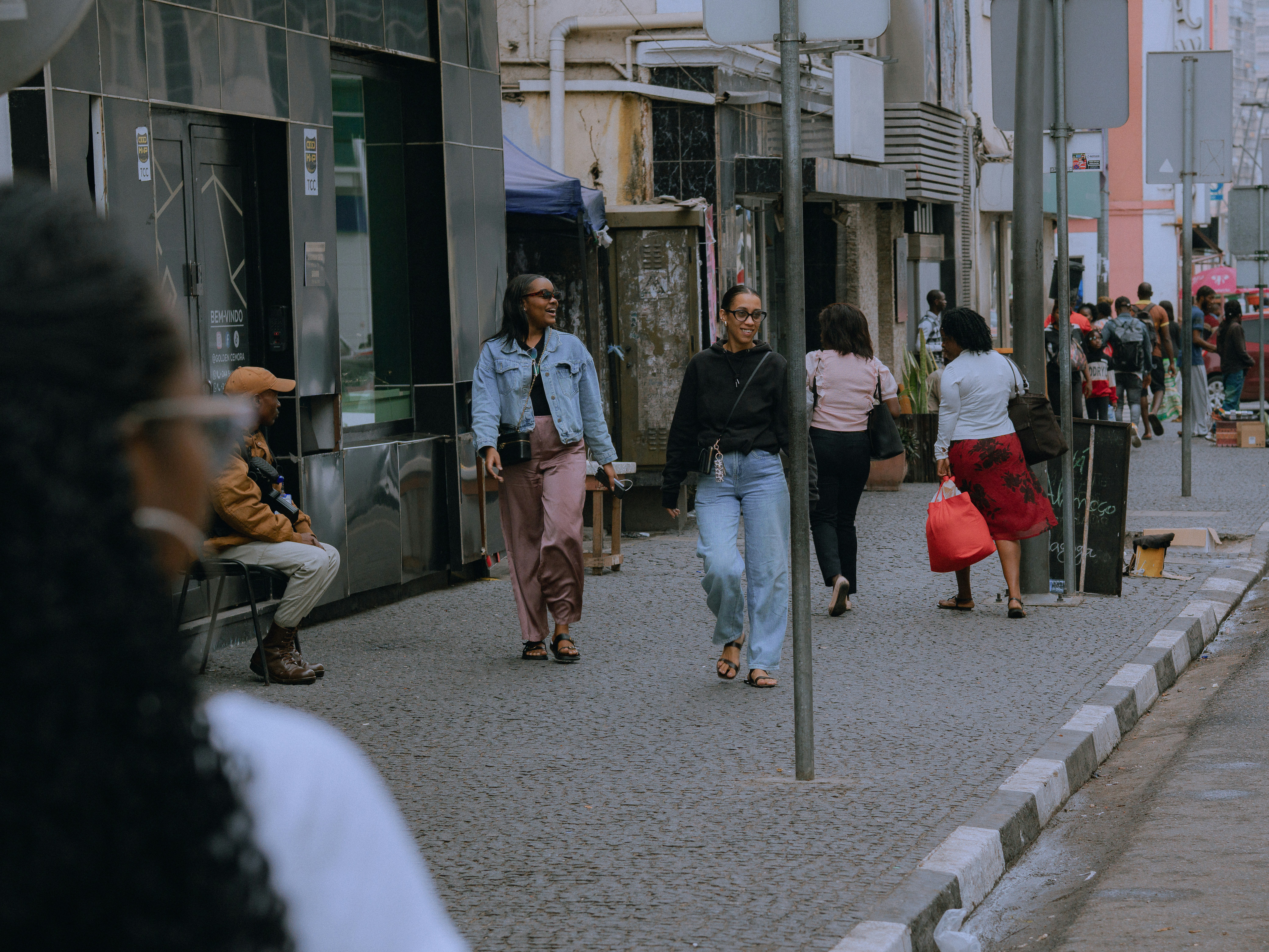 Two women stroll along a bustling street, surrounded by various pedestrians and storefronts, capturing the essence of urban life.