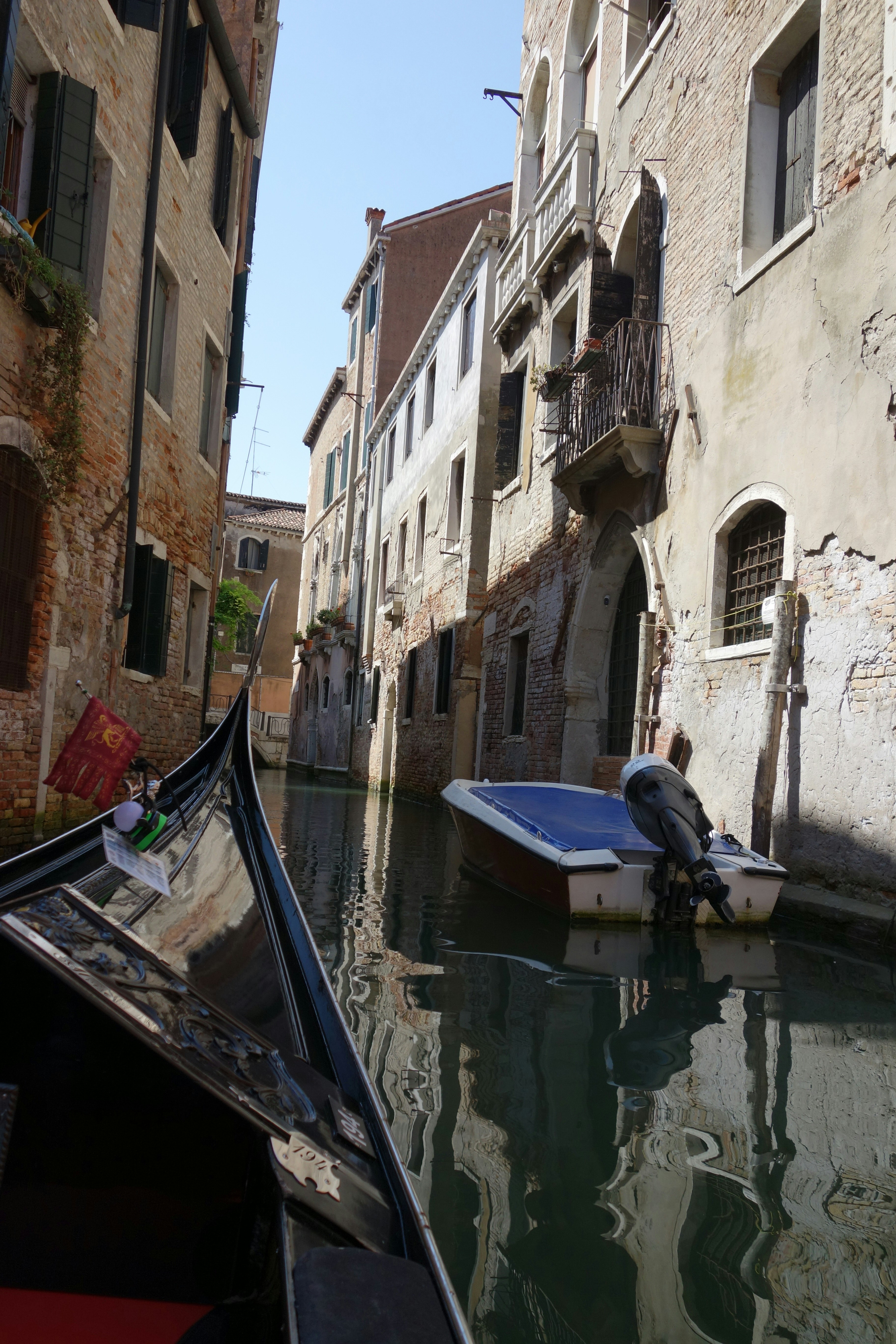 Narrow canal flanked by weathered buildings, with a gondola in the foreground and a small boat moored nearby. The scene captures the tranquil essence of Venetian waterways.