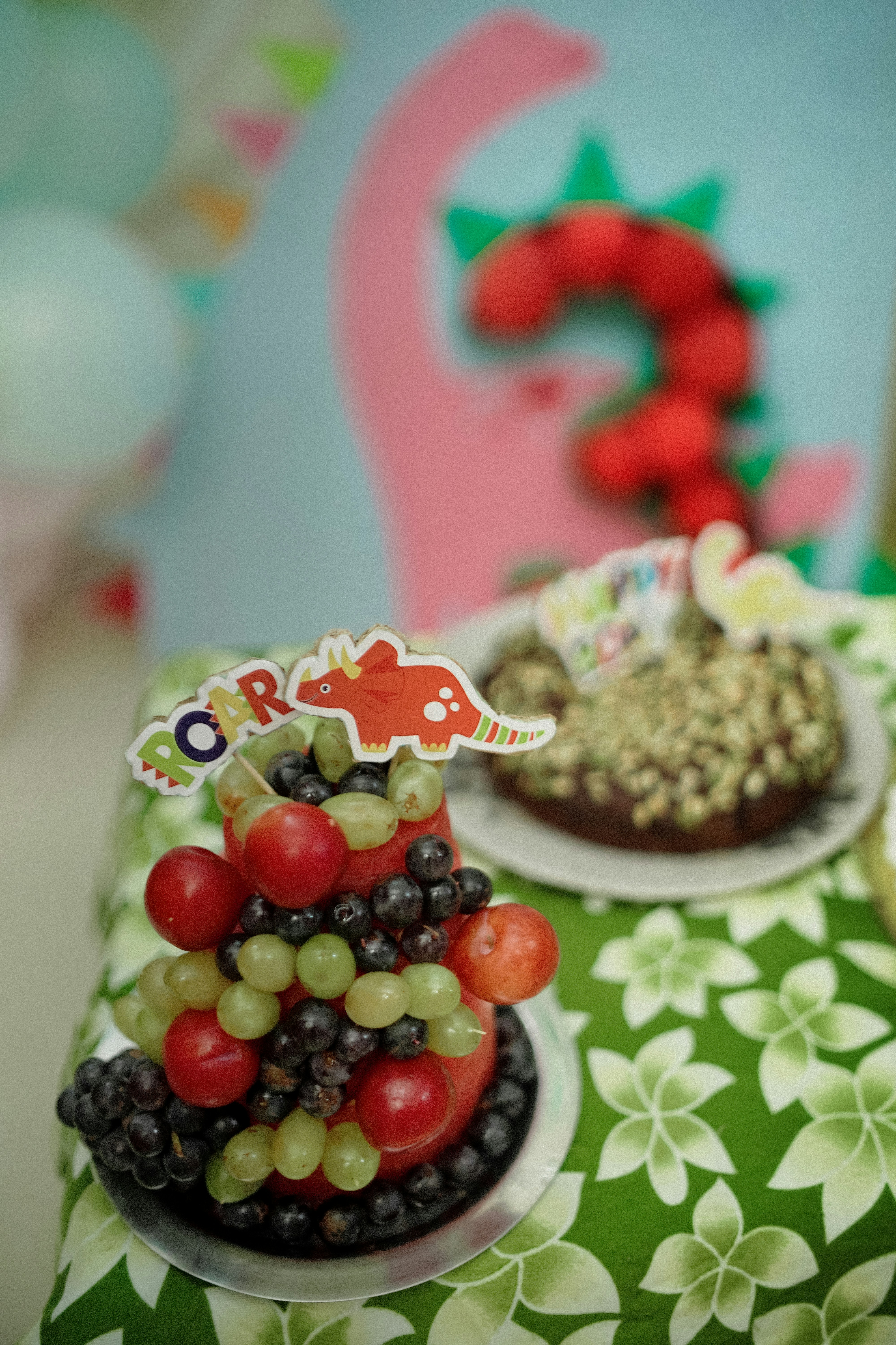 These photos capture a fun and sweet dinosaur-themed third birthday party. The vibrant decor features a balloon arch in soft pastel colors, a large cutout of a pink dinosaur, and a "3" decorated with red balloons. The birthday girl, dressed in a "Roar, I'm 3" t-shirt, is seen posing with her family and enjoying her special day. The collection includes shots of the festive setup, delicious-looking birthday cakes, and the pure joy of the celebration. | Fruit tower and donut at a dinosaur themed party.