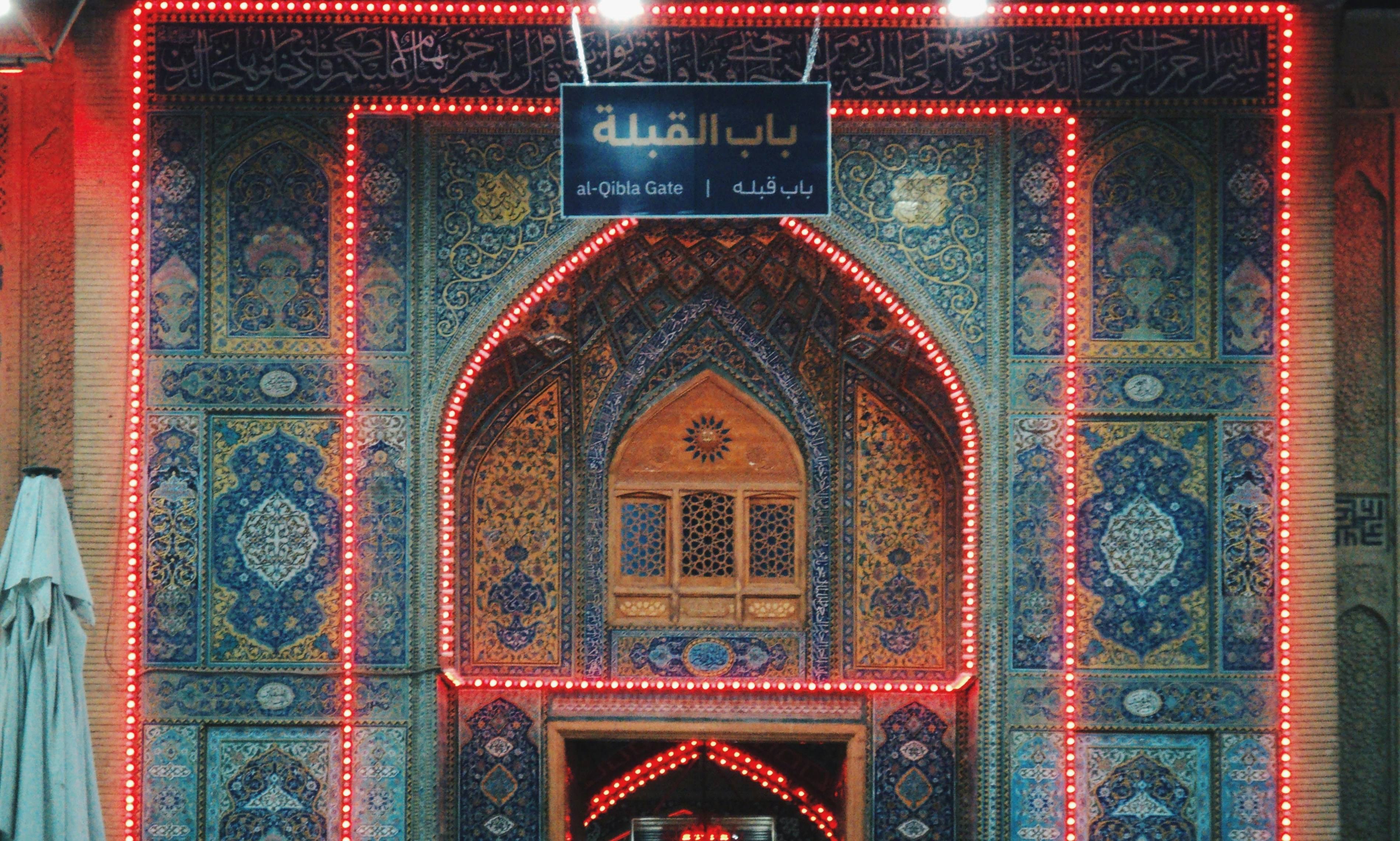 Ornate entrance of the Al-Qibla Gate adorned with vibrant blue tiles and illuminated by red lights, showcasing intricate designs and cultural significance.