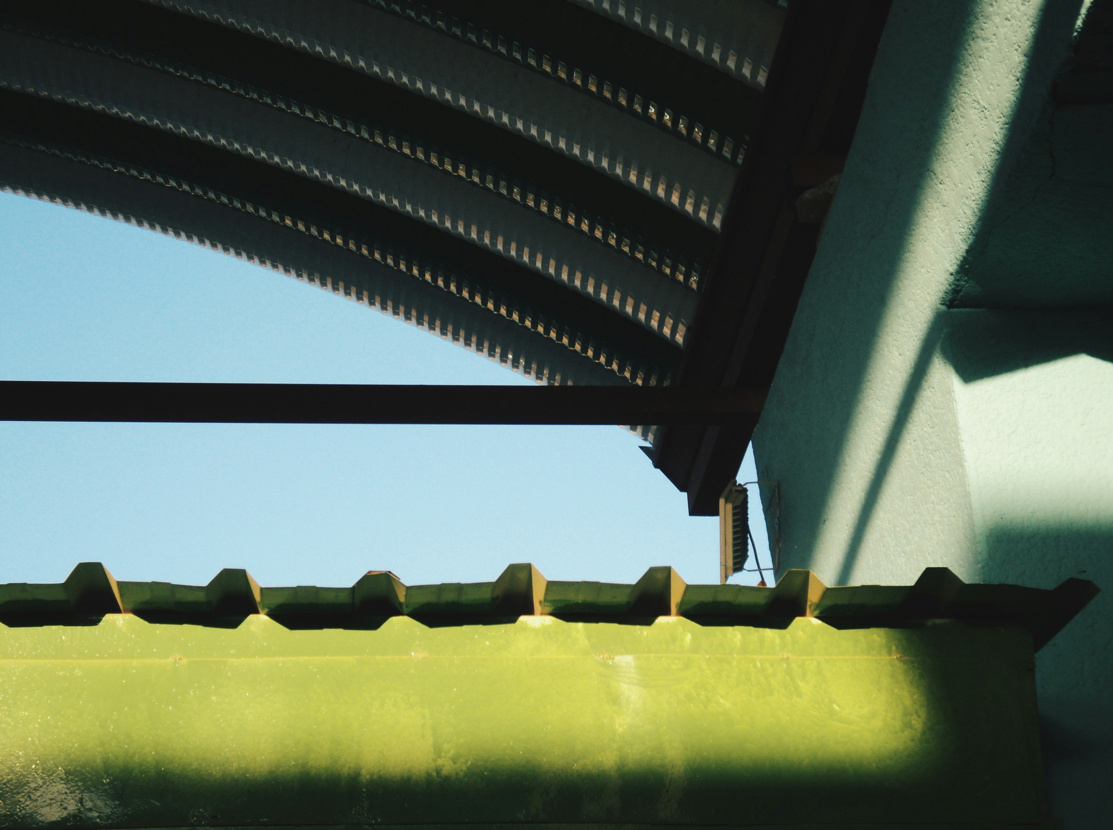 Abstract composition featuring a green rooftop under a corrugated metal structure with a clear blue sky above.