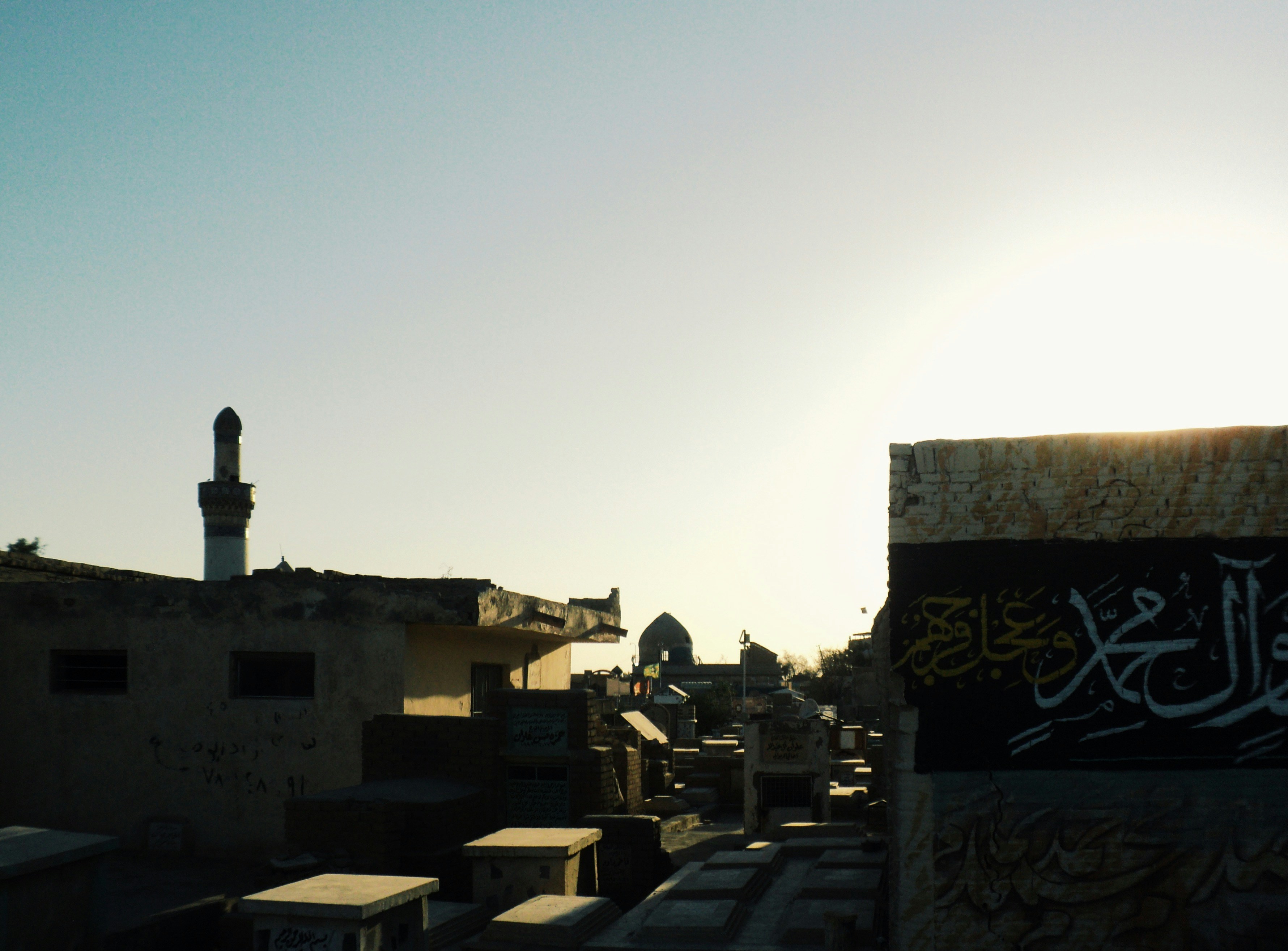 A serene cemetery scene at dusk, featuring intricately designed tombstones with Arabic calligraphy against a backdrop of distant minarets and a fading sun.