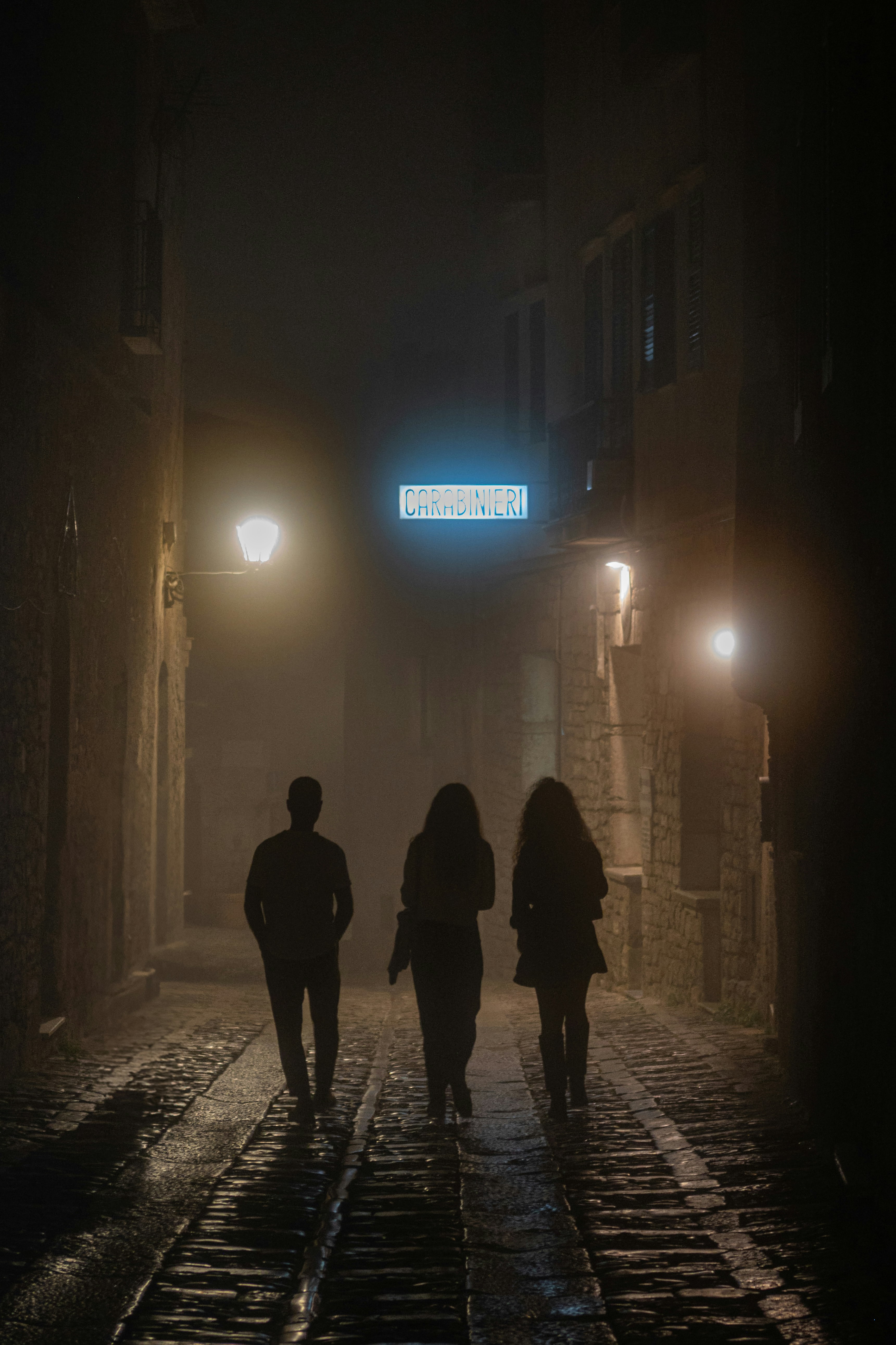 Three figures walk down a foggy cobblestone street at night.