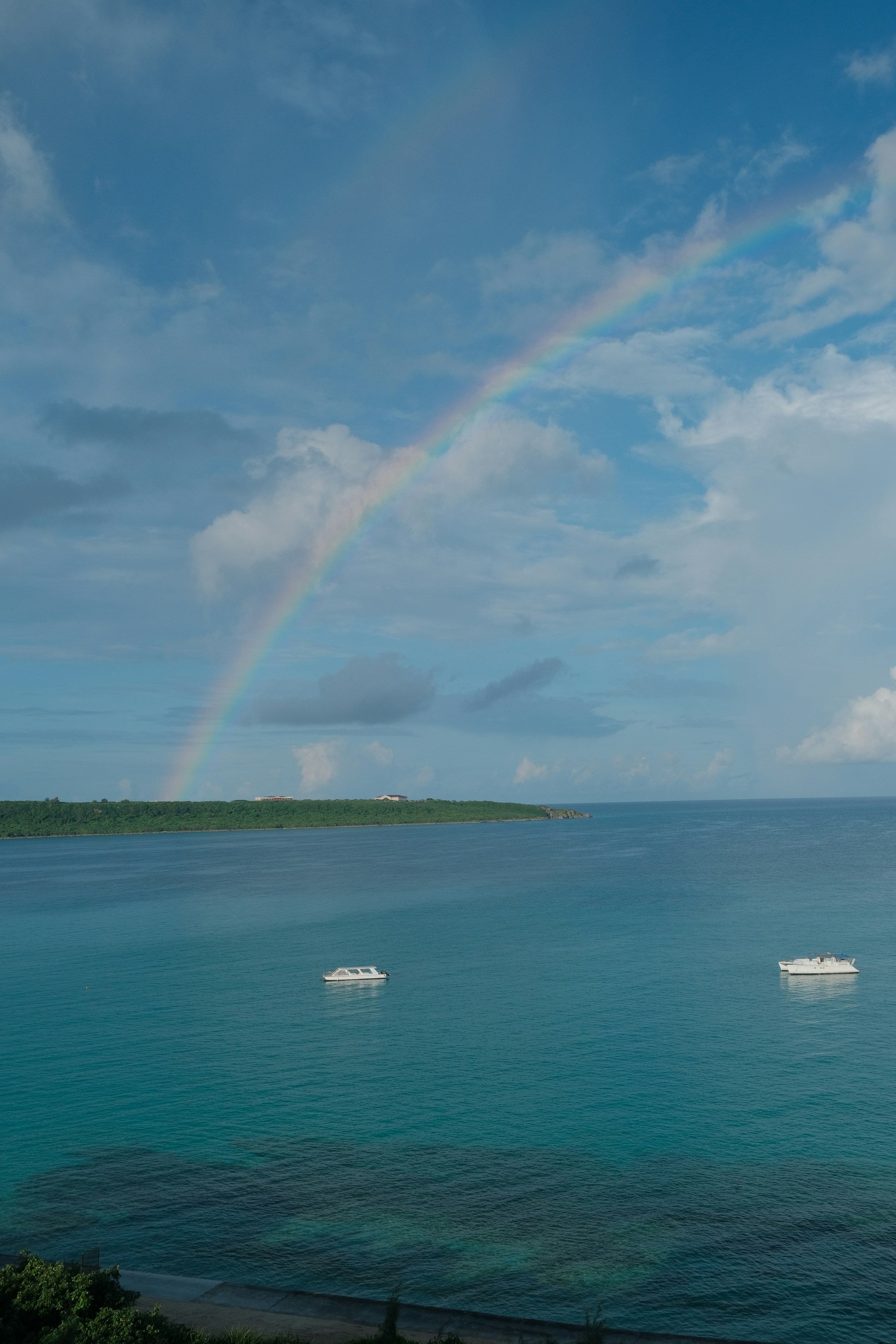 A vibrant rainbow arches over a tranquil sea, with boats gently bobbing on the turquoise waters near a lush green shore.