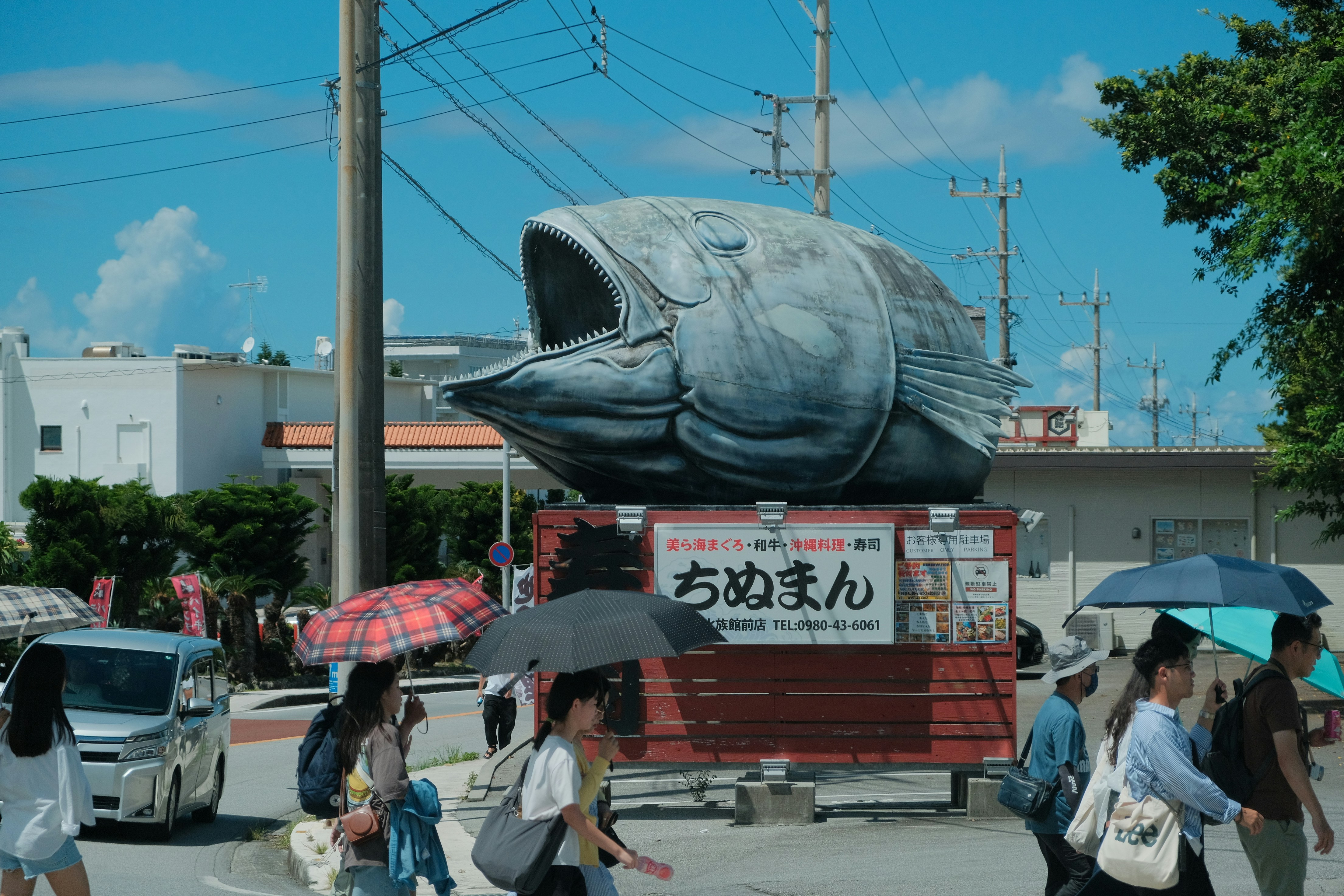 A large fish sculpture dominates the scene, surrounded by pedestrians and vehicles on a sunny day. The vibrant blue sky adds to the lively atmosphere.