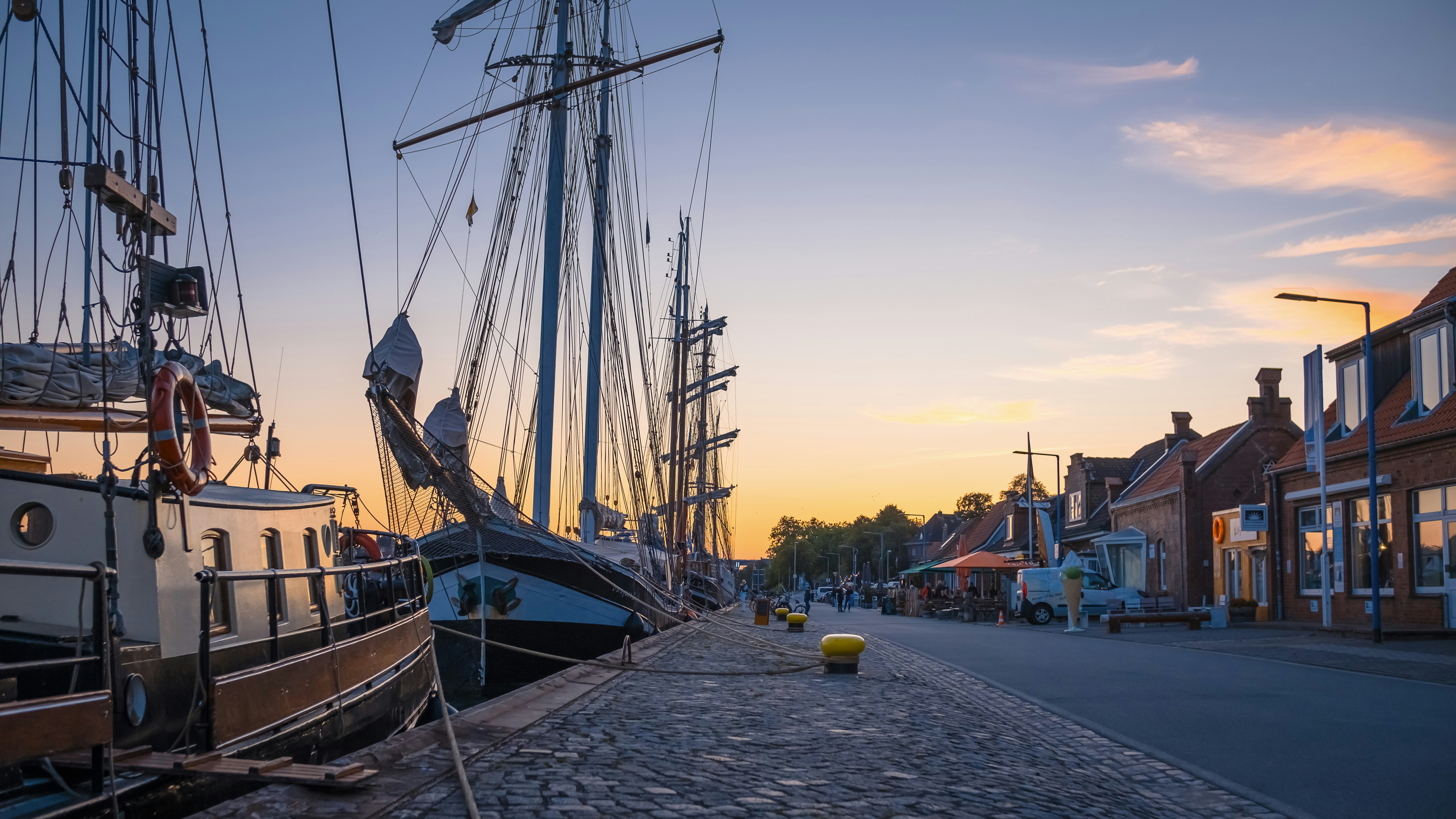 Sailing boats lined up at a tranquil harbor during sunset, with quaint buildings in the background. 
