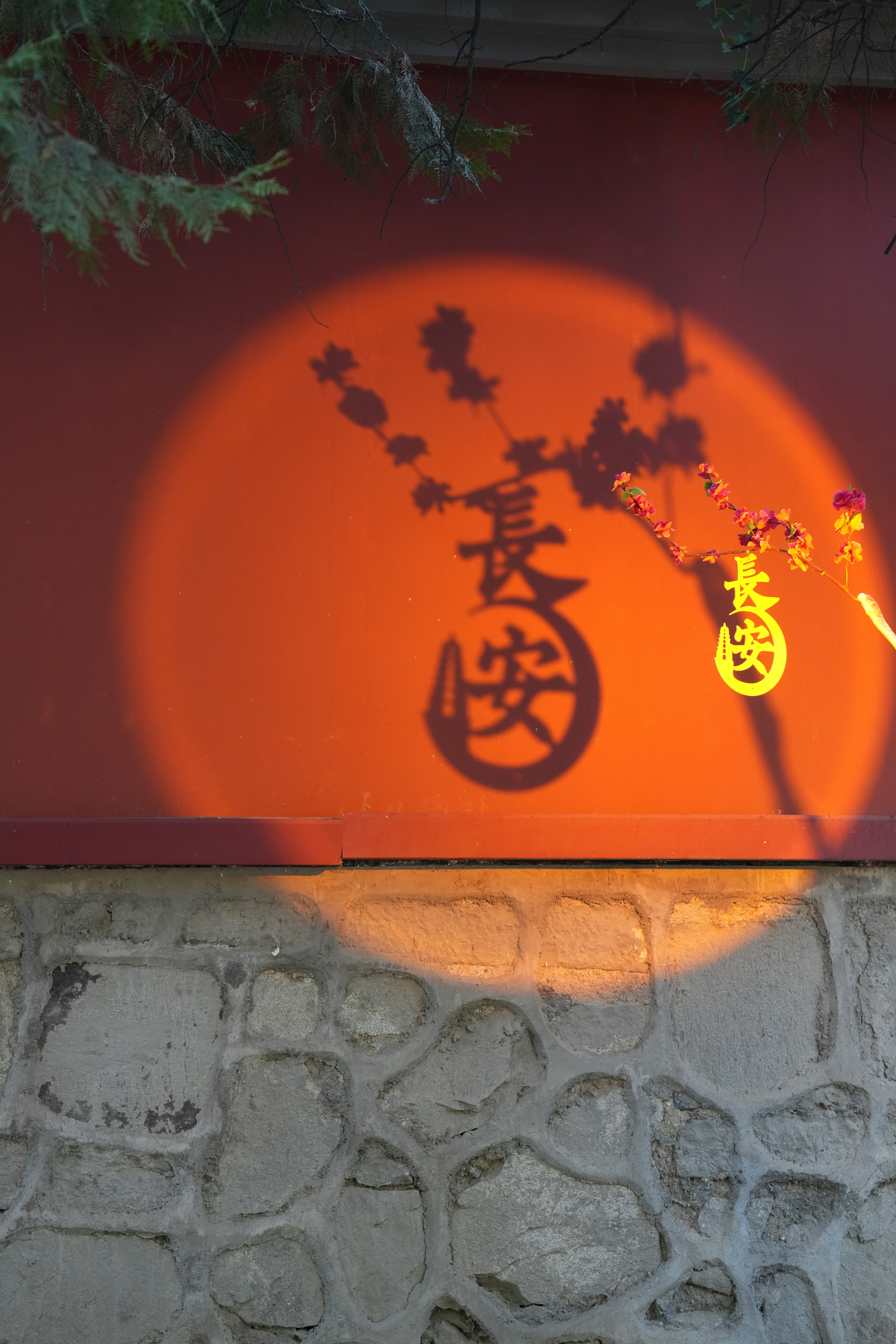 Red wall with a circular light highlight casting a dark calligraphic shadow and a hanging ornament against a stone base.