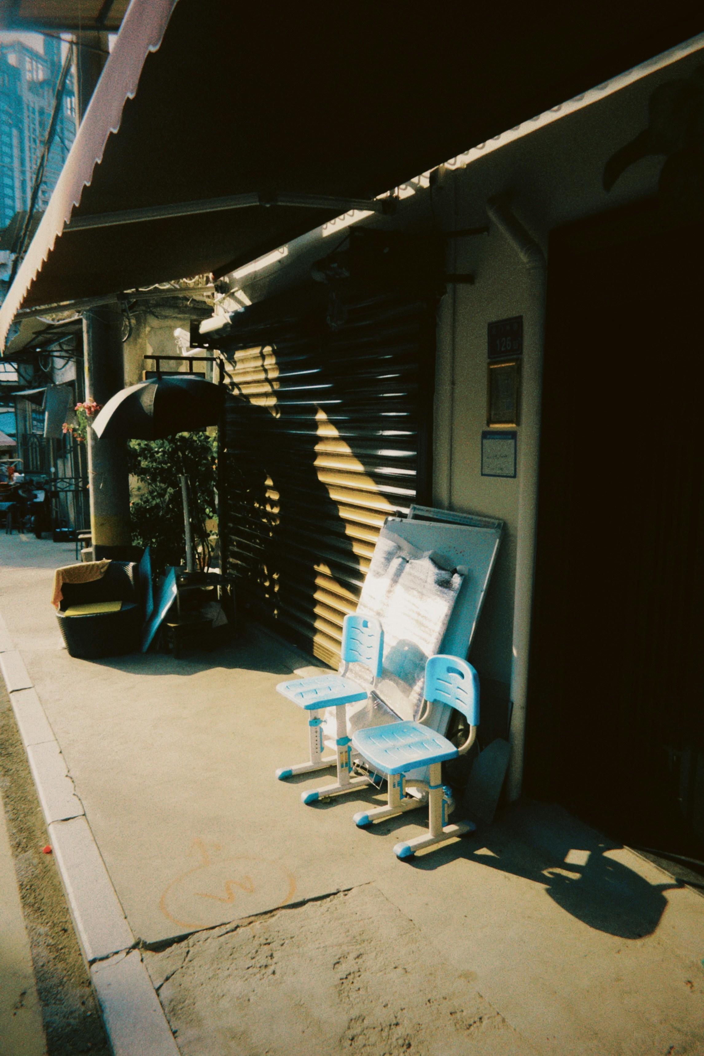 Two blue plastic chairs sit abandoned beside a shadowy entrance, hinting at stories of the past. The scene captures a moment of stillness in a bustling urban environment.