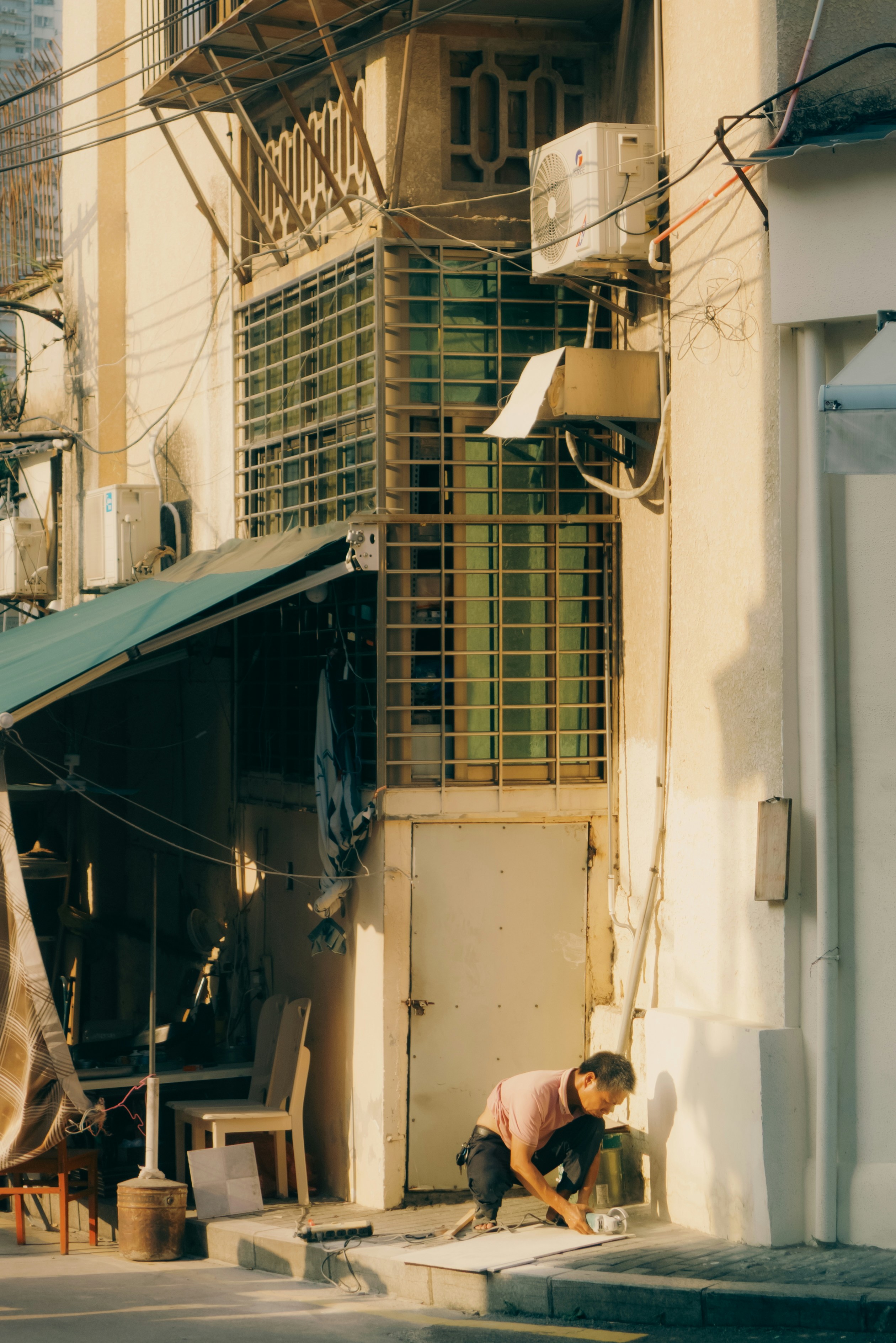 A man kneeling on the sidewalk, focused on his task amidst a backdrop of urban architecture and tangled wires. The scene captures the essence of everyday life in a bustling city.