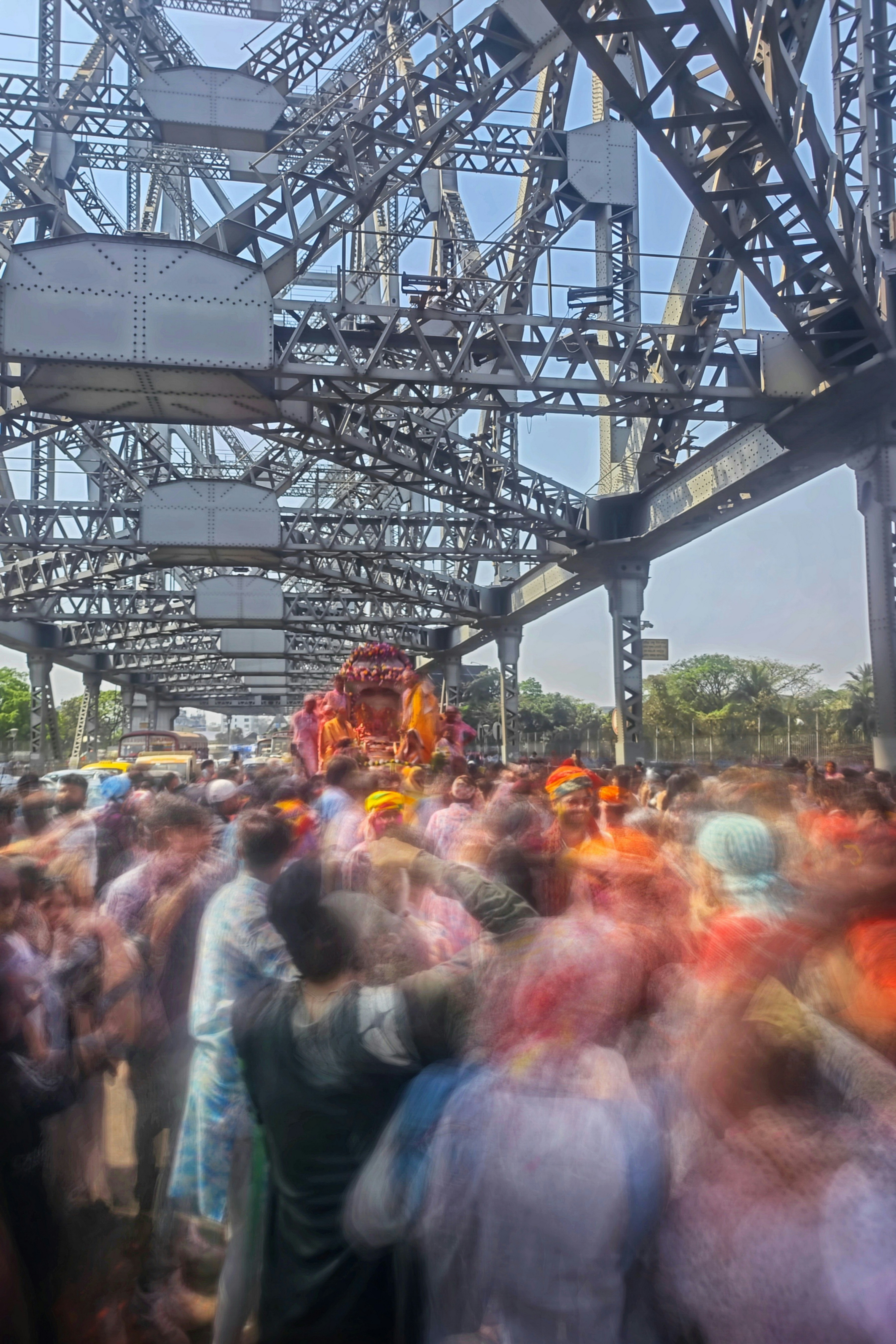 Vibrant festival scene with a crowd celebrating beneath a grand iron bridge, showcasing colorful attire and joyful expressions.